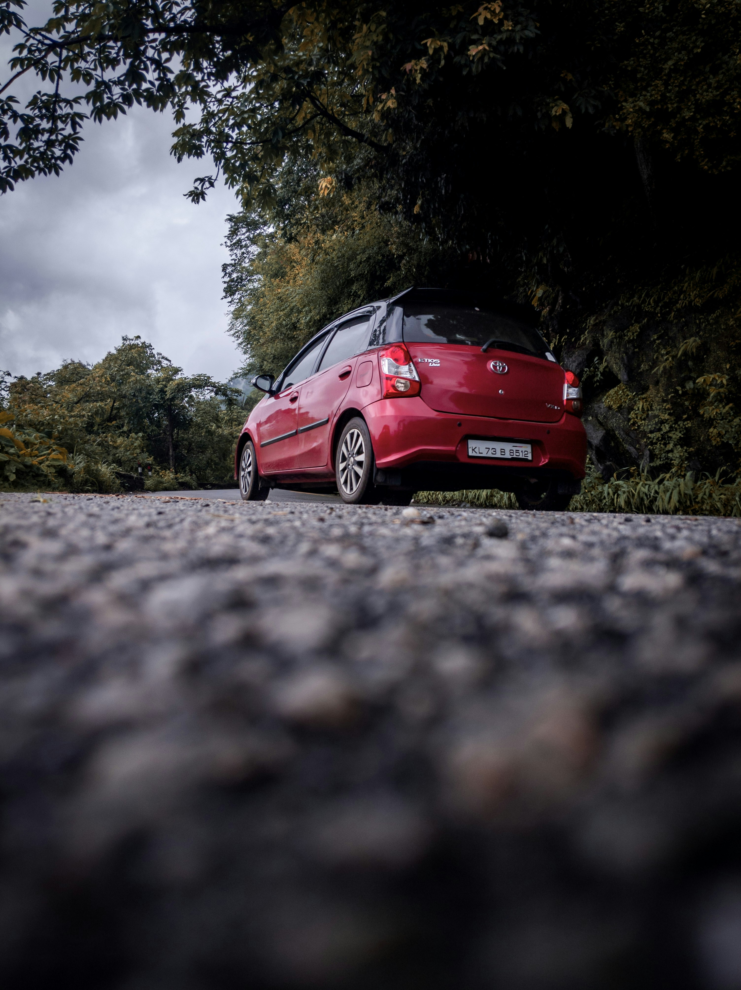 Red hatchback under a leafy overhang, parked on a damp gravel road
