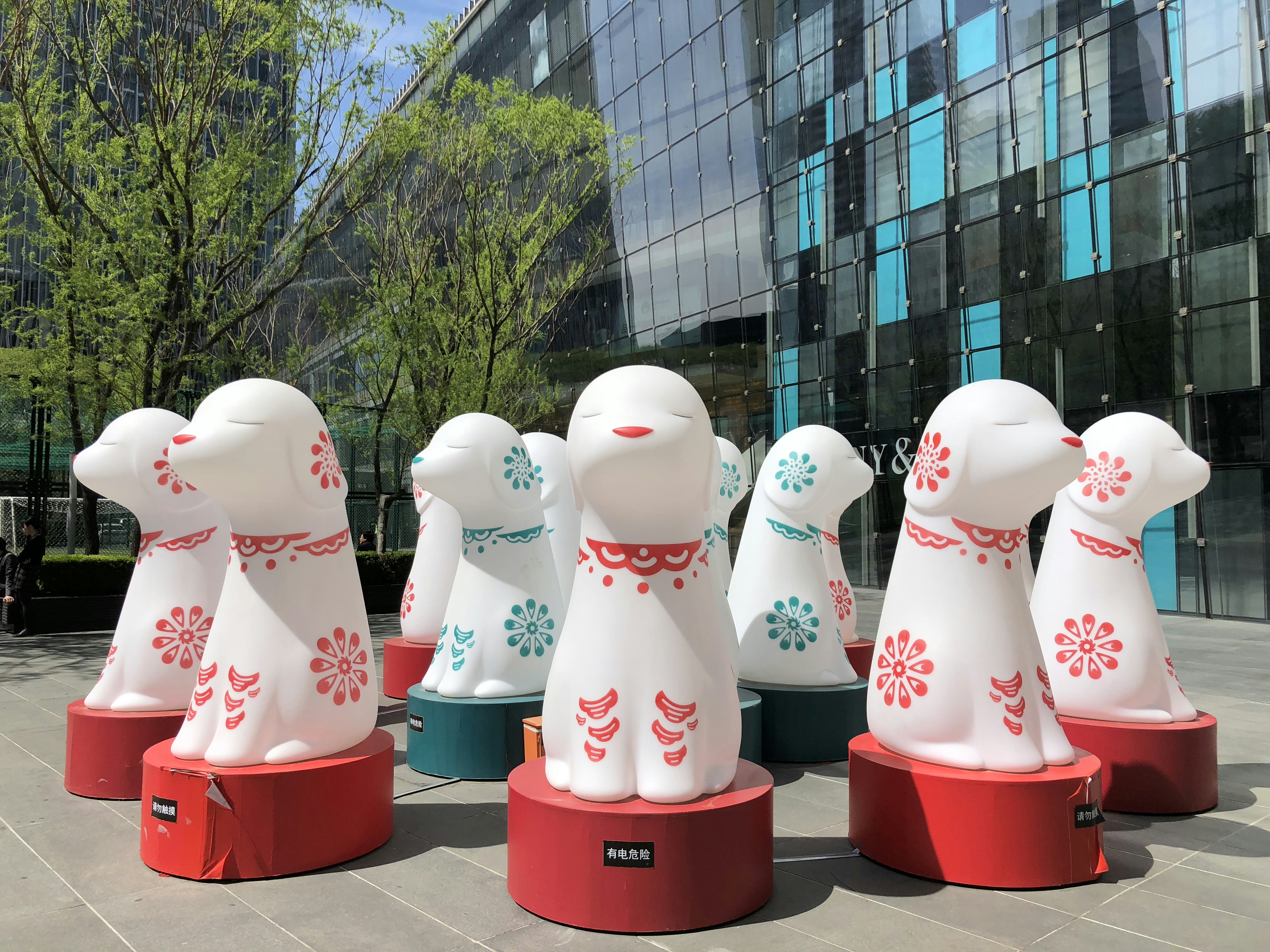 Colorful, oversized dog sculptures arranged in a playful formation outside a modern building, surrounded by greenery.