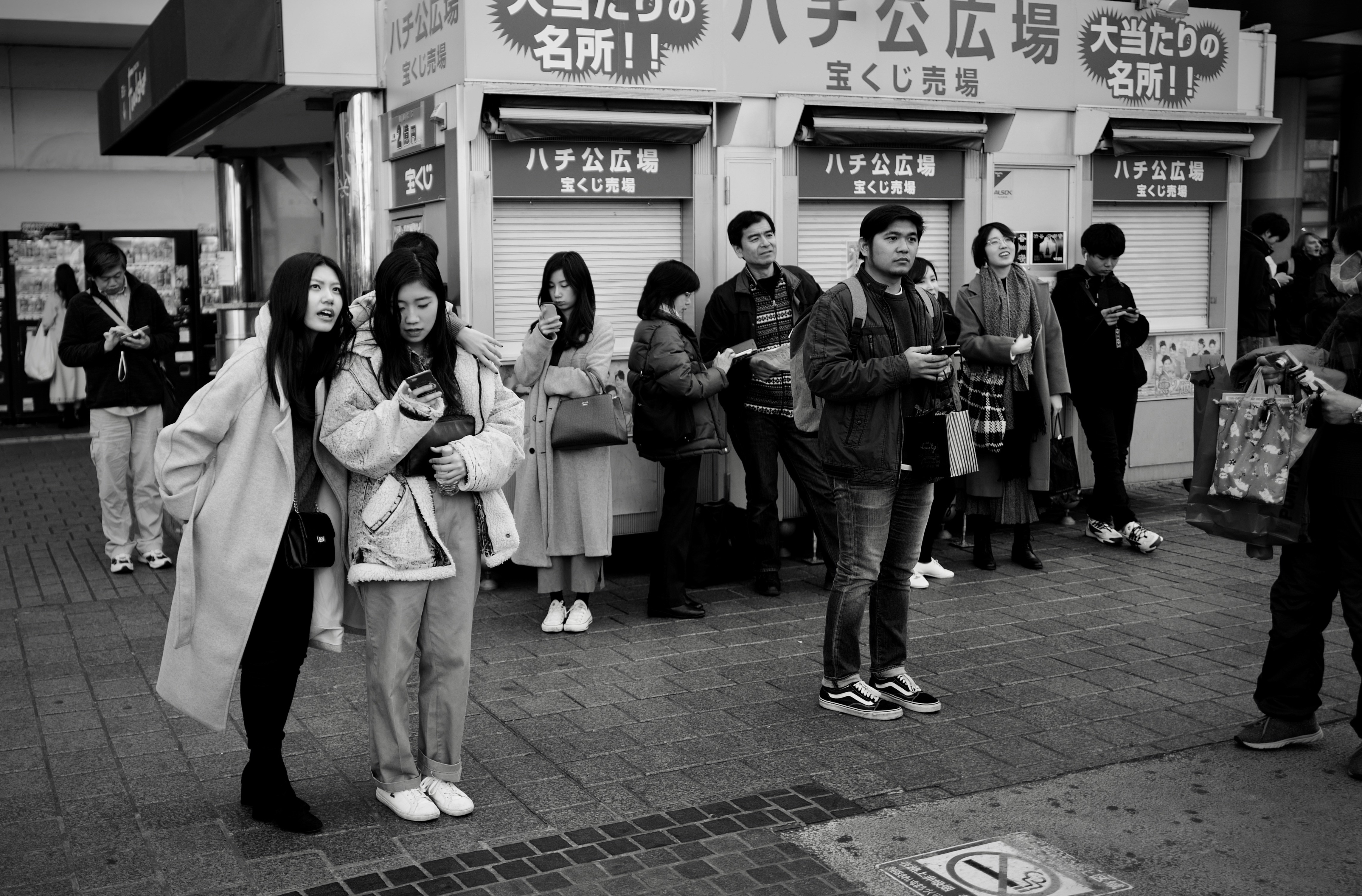 Two friends share a lighthearted moment while others are absorbed in their phones in a bustling urban setting.