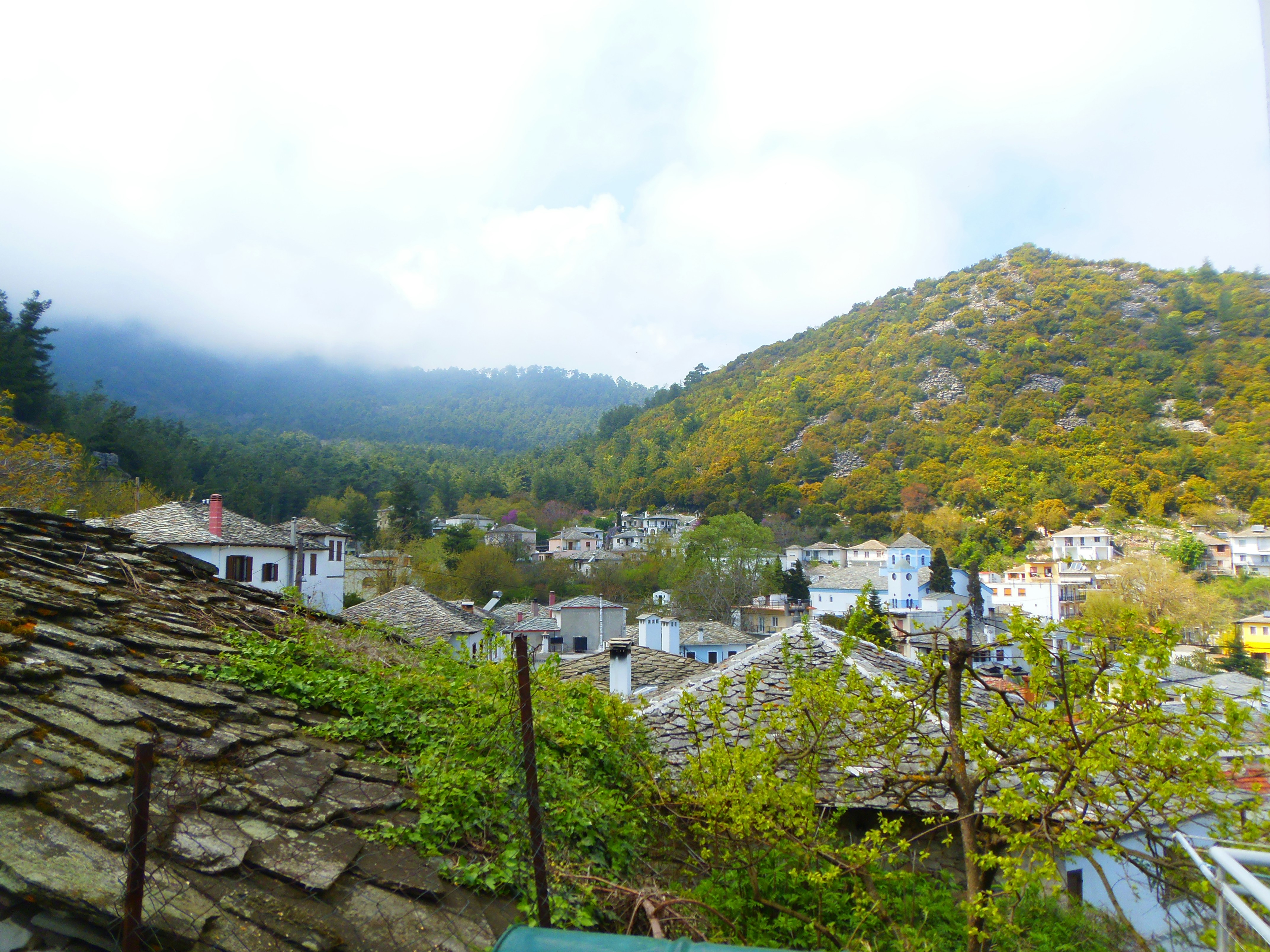 Lush green hills cradle a quaint village nestled among rustic rooftops, with a hint of fog enveloping the mountainside.
