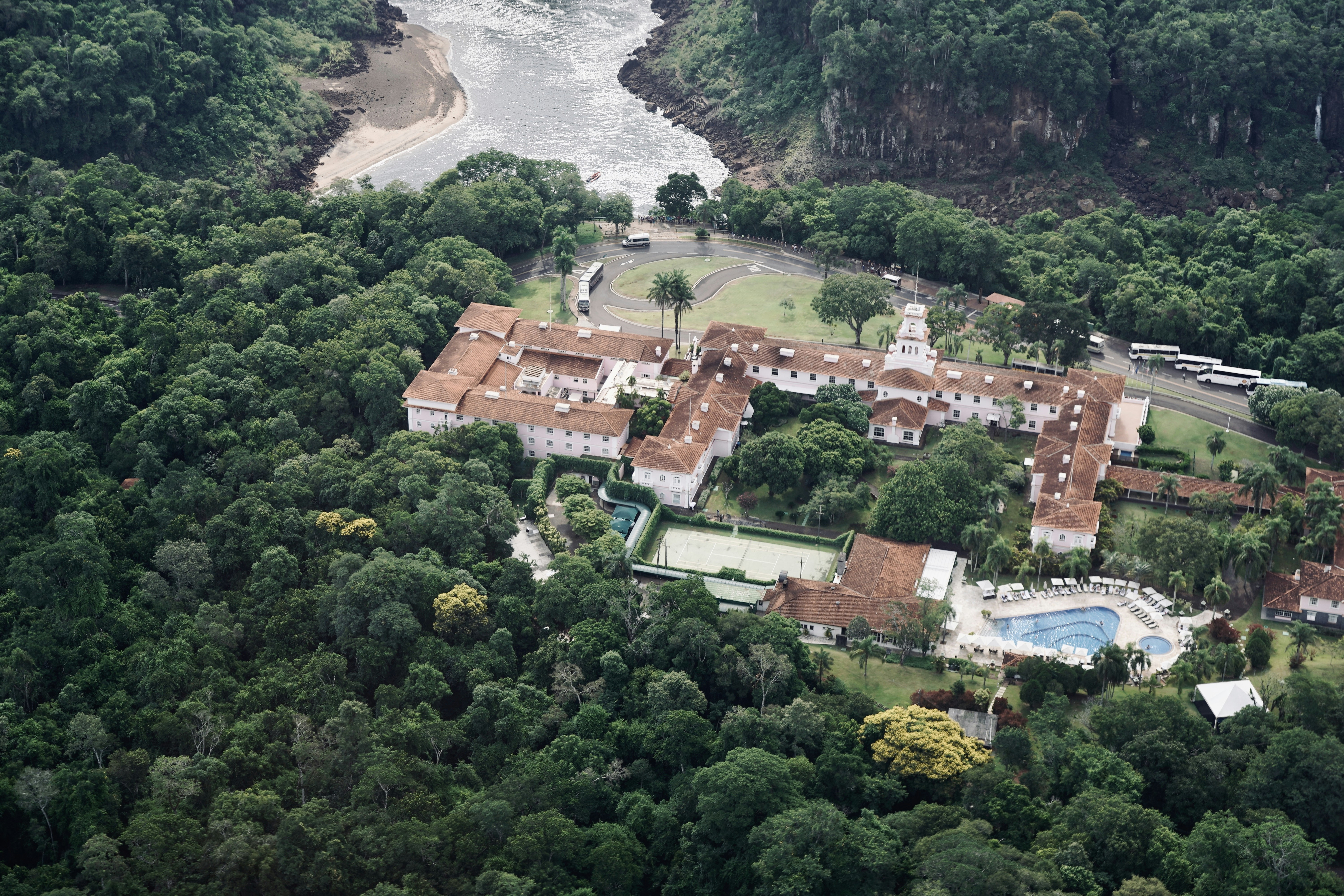 Aerial view of a sprawling complex with red roofs nestled in dense green forest near a winding river.