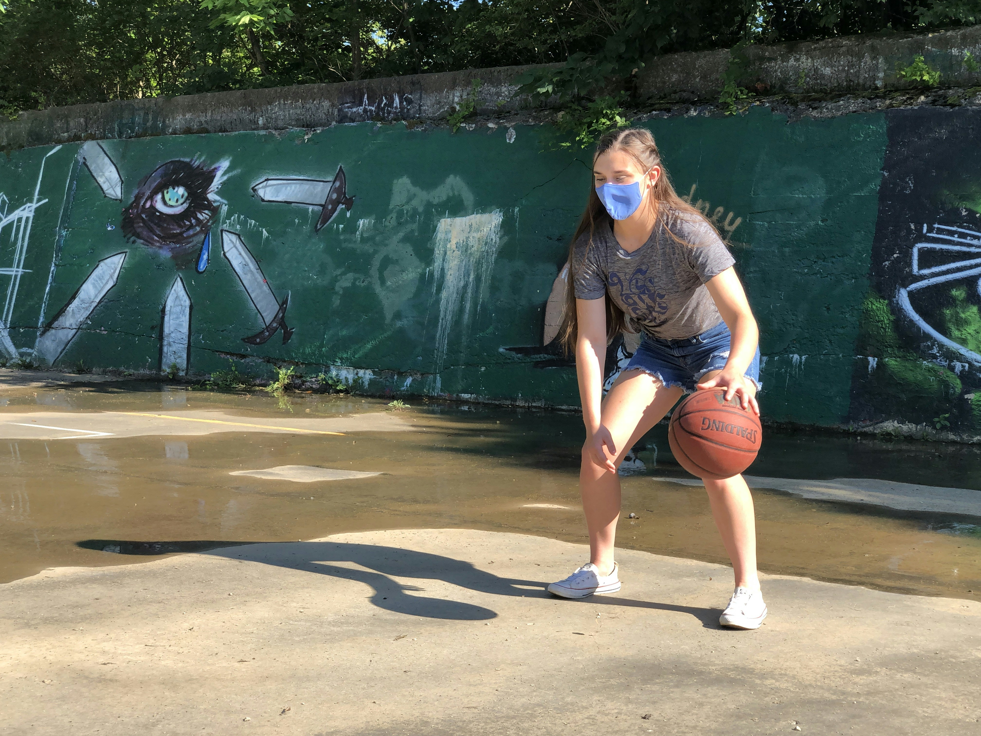 Young woman in a blue mask prepares to dribble a basketball on a sunlit urban court, with vibrant graffiti in the background.