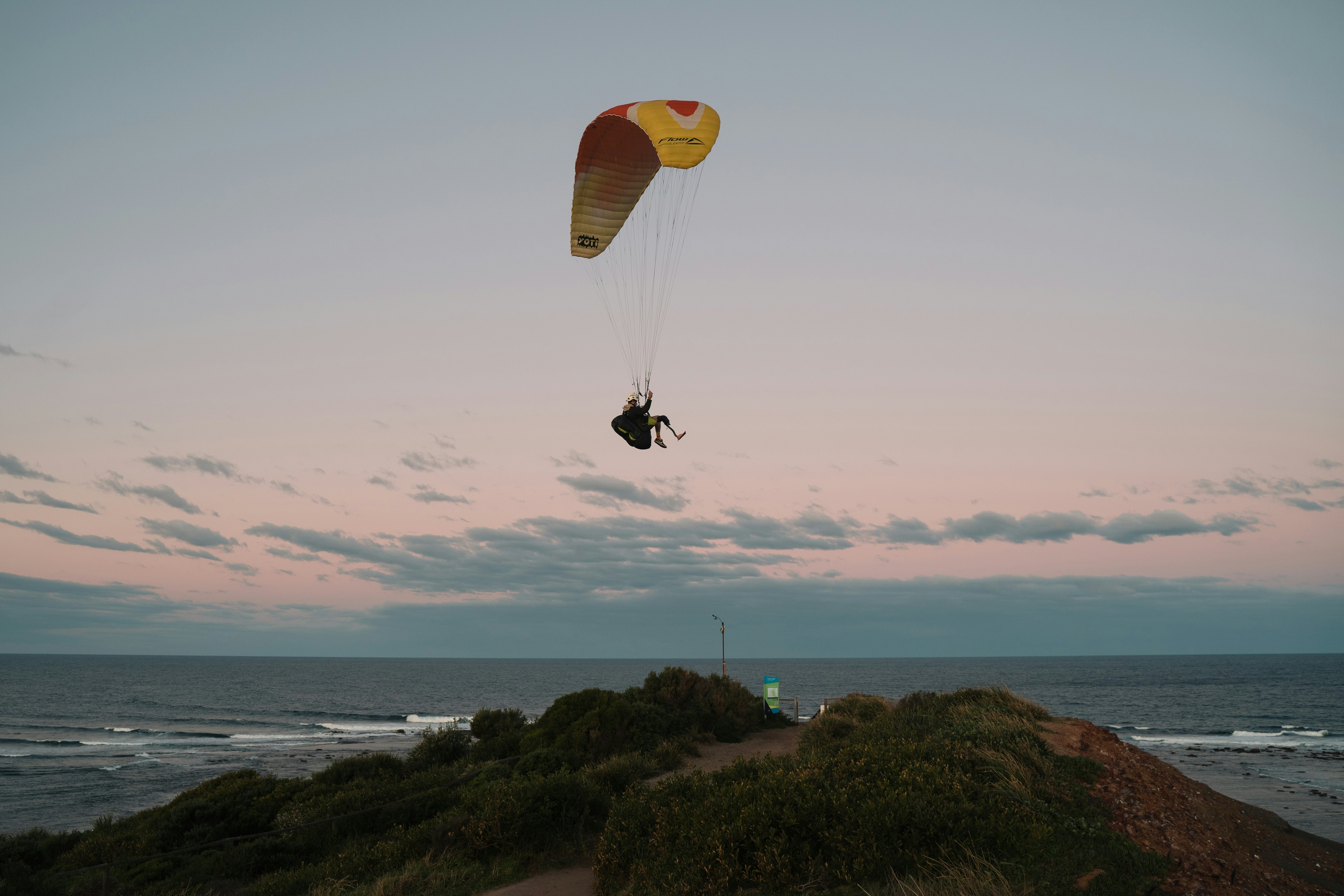 A paraglider gracefully descends over a coastal landscape at dusk, with gentle waves lapping at the shore below. The sky transitions from soft pink to deep blue.