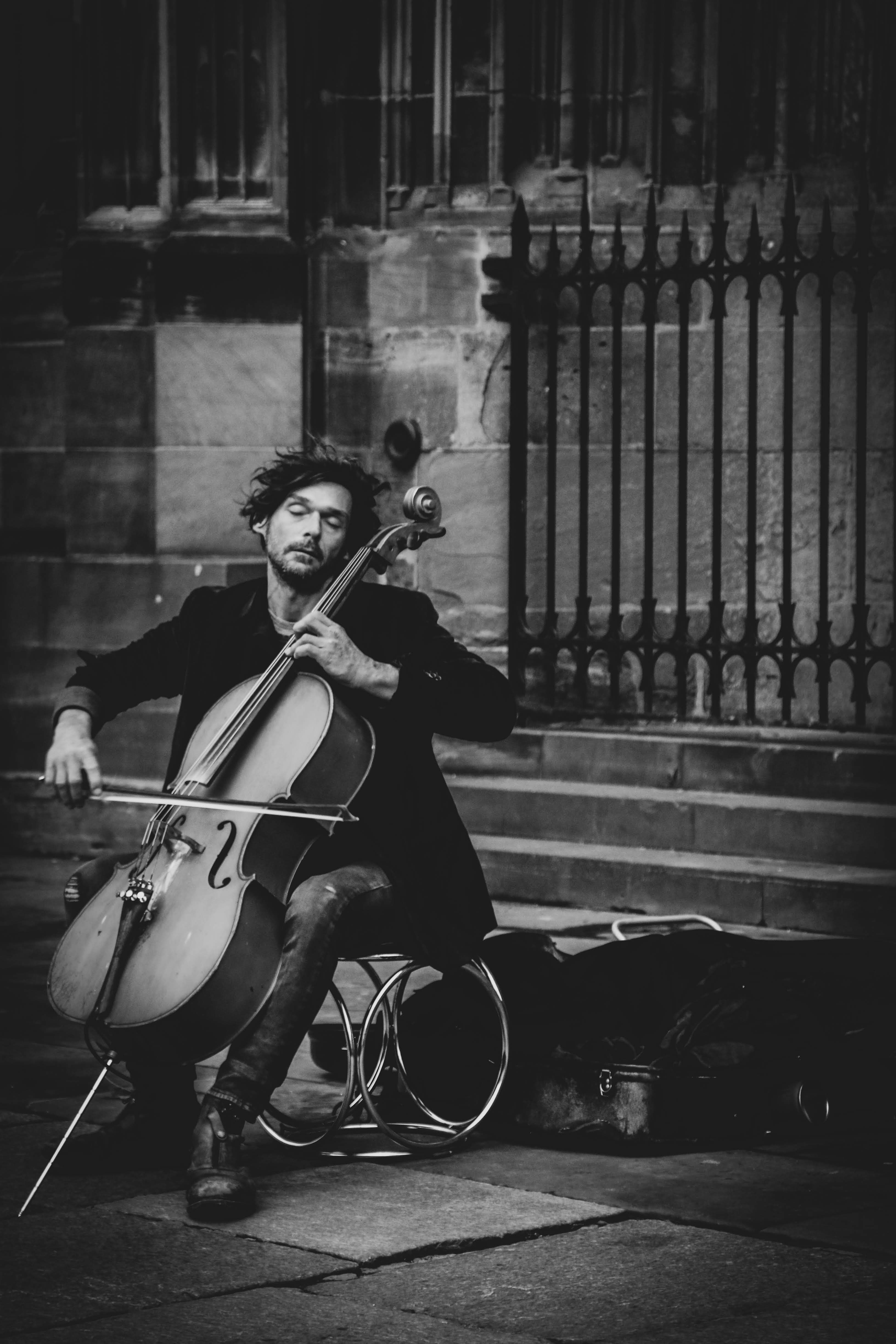 A cellist immersed in his music, seated against a textured stone backdrop with a vintage suitcase nearby. The monochrome tones enhance the emotional depth of the scene.