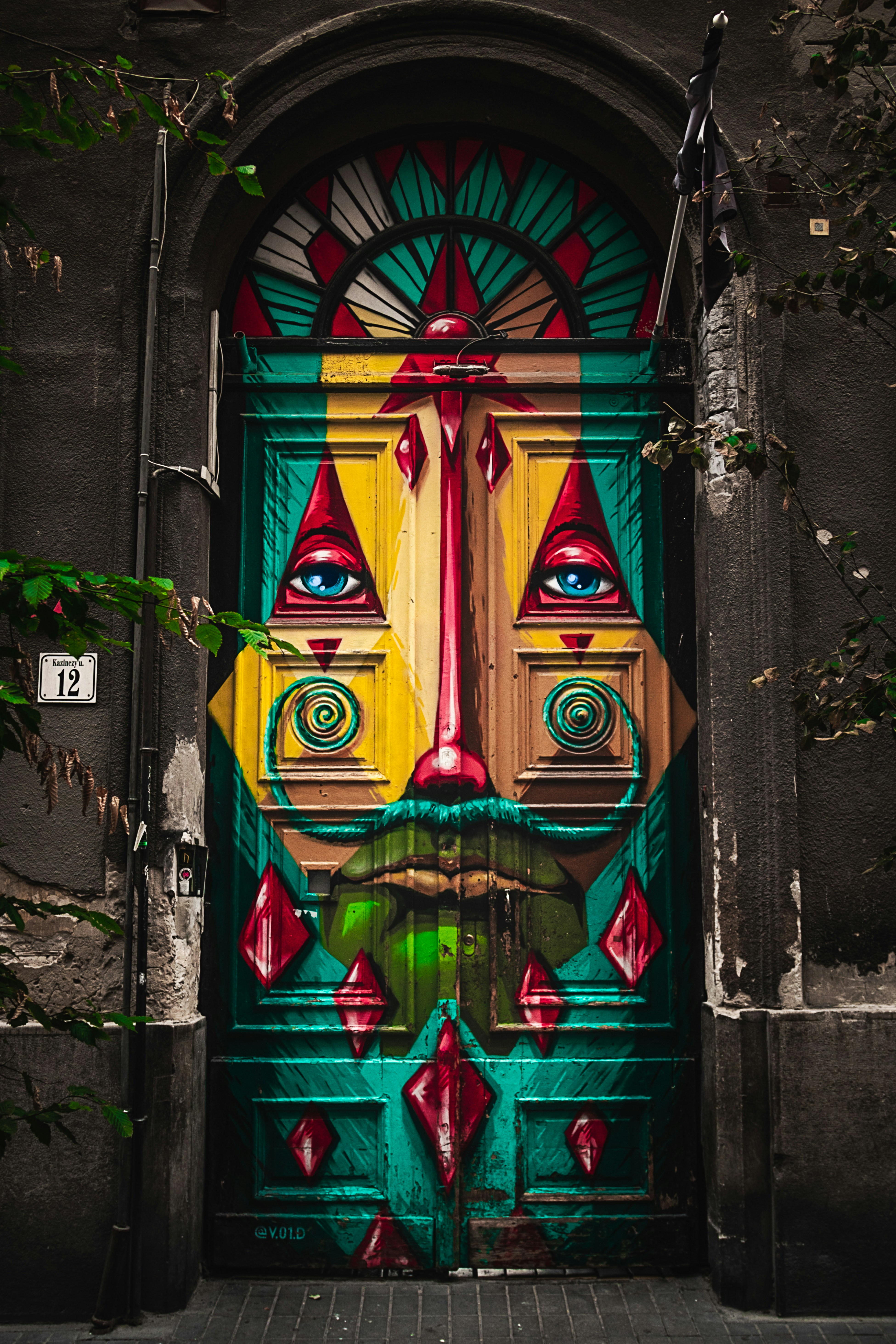 Vibrant mural depicting a stylized face on a wooden door, surrounded by urban foliage. The artwork features geometric shapes and bold colors.