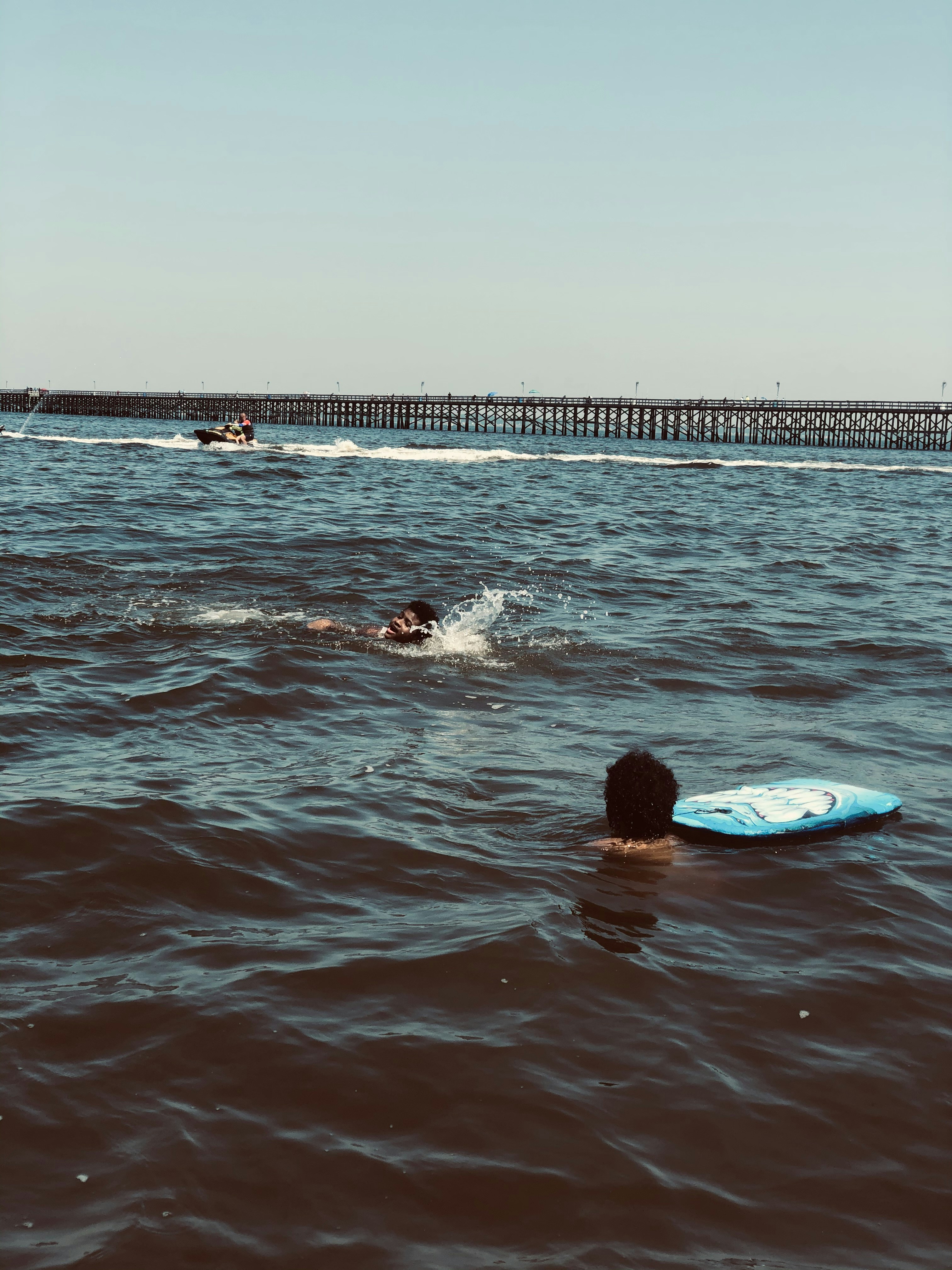 Children swimming in the ocean near a long pier under a clear sky.