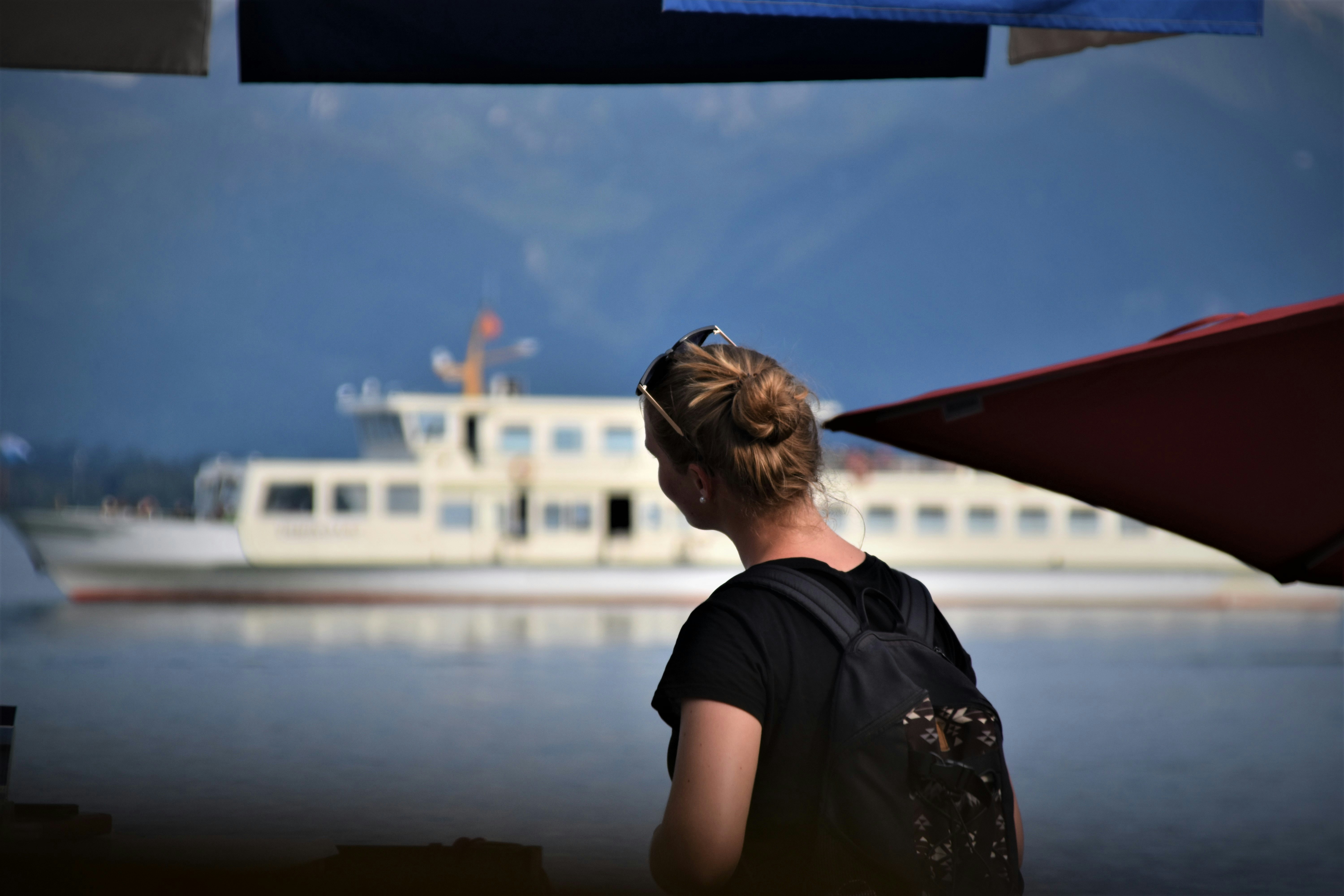 A woman gazes thoughtfully towards a distant boat, framed by colorful canopies and mountains in the background.