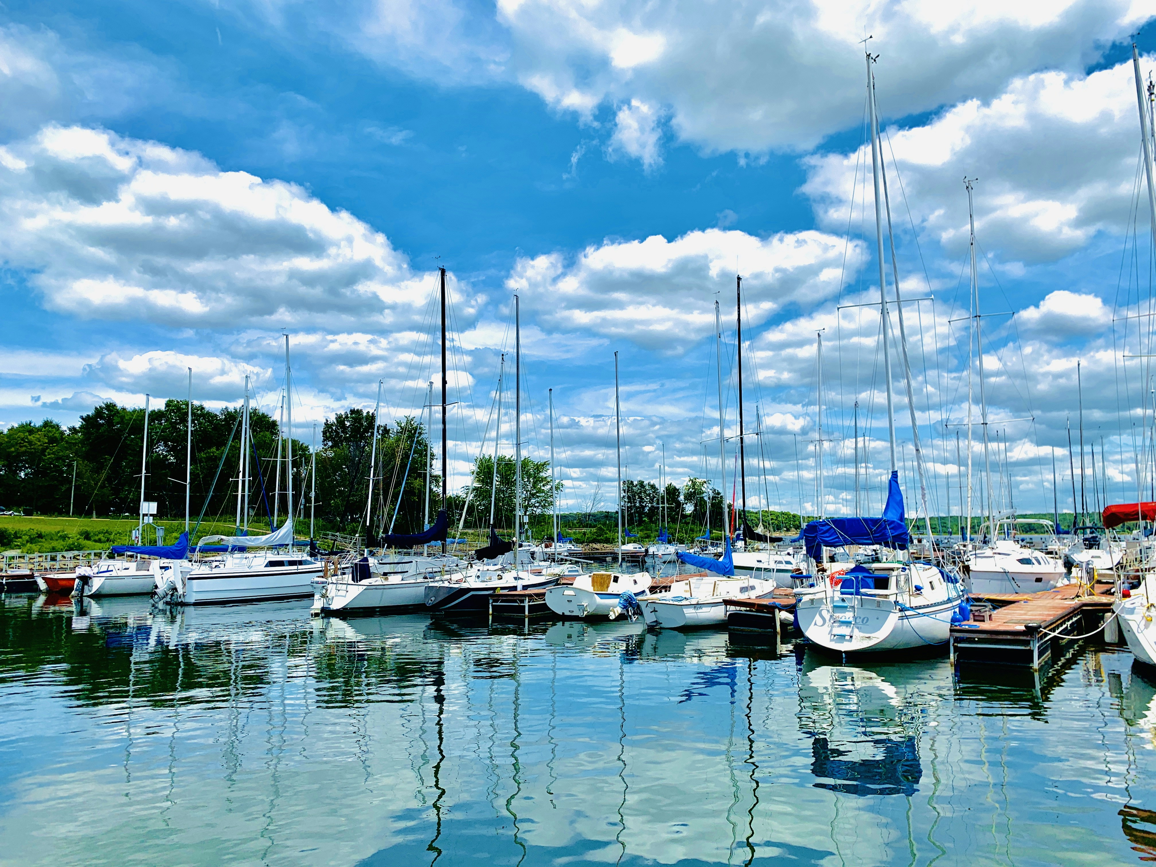 Sailboats moored in a marina under a vibrant blue sky with fluffy clouds reflected in calm water.
