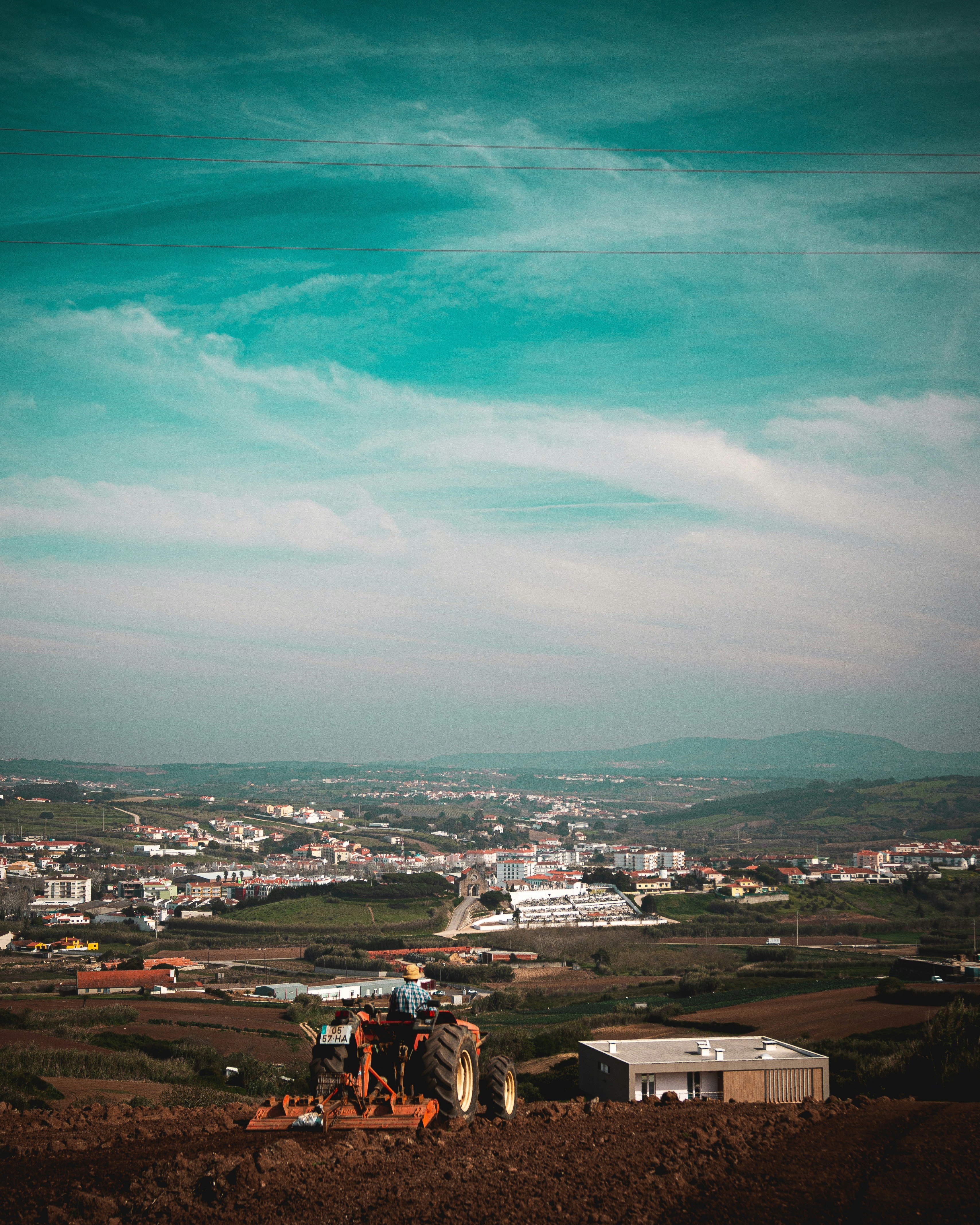 Tractor plowing a field with a sprawling town and hills in the background under a vibrant sky.