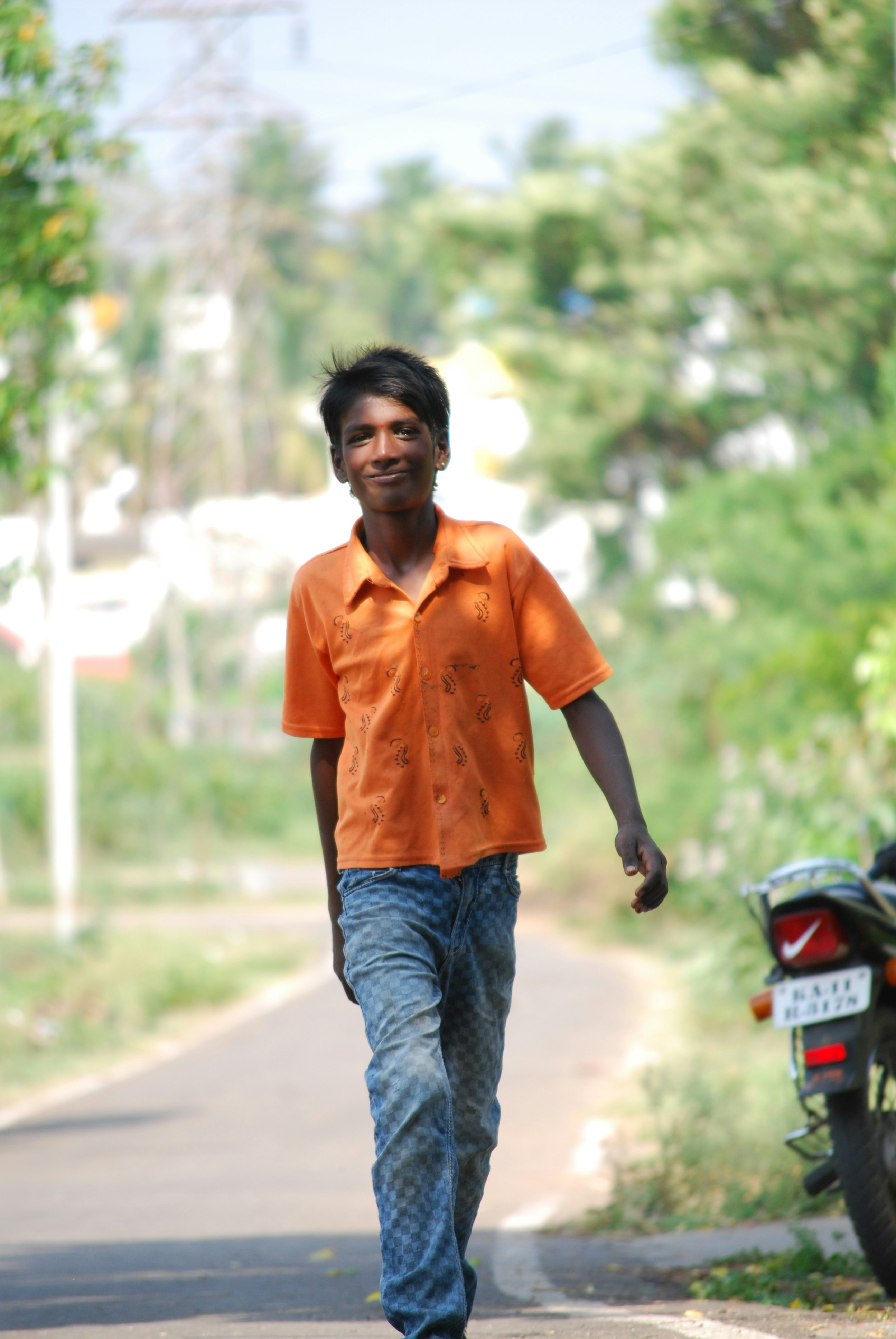 A young boy in an orange shirt walks confidently along a tree-lined street, showcasing a moment of carefree joy.