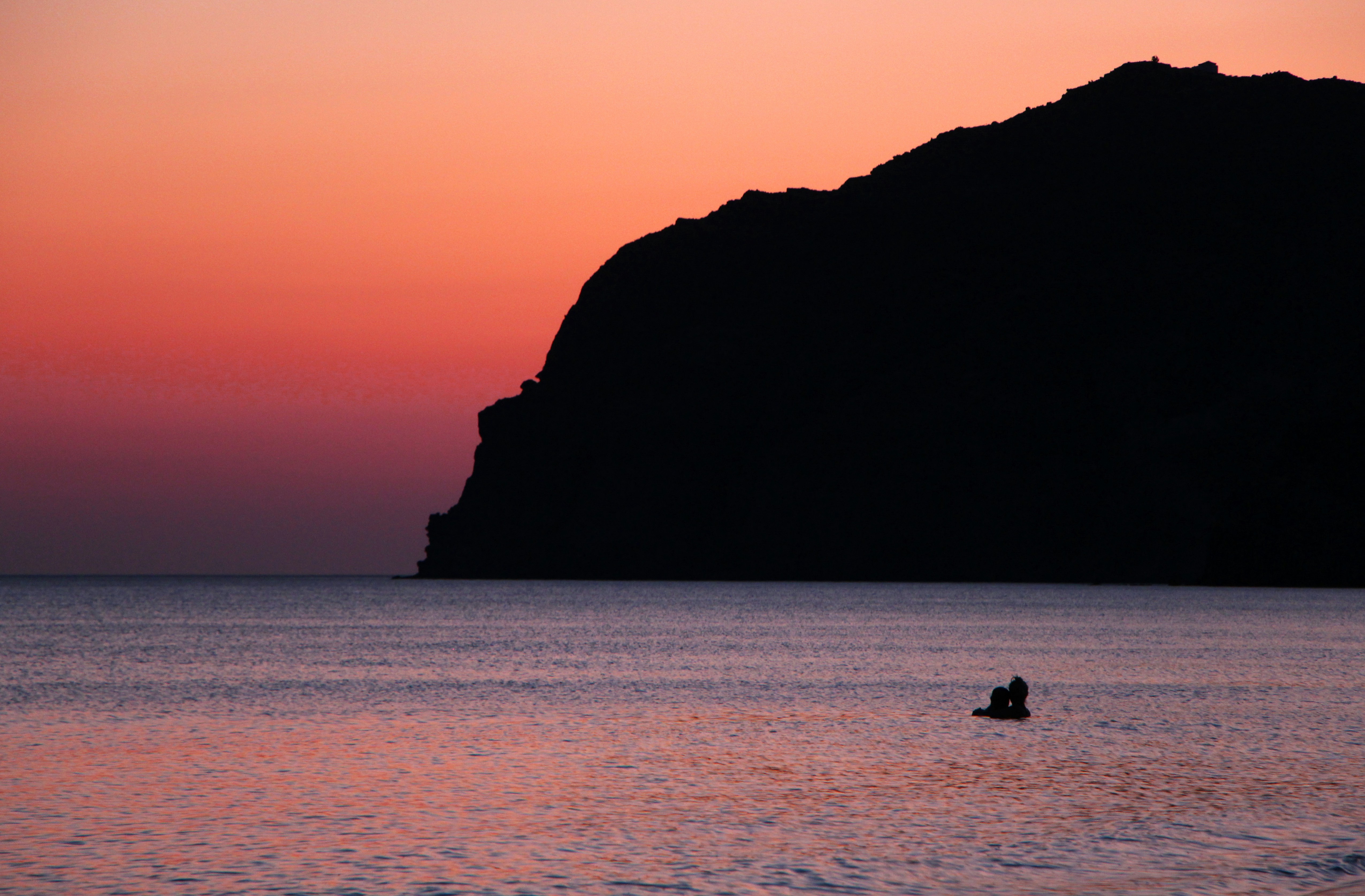 A couple silhouetted against a vibrant sunset, with a rocky coastline in the background. The calm water reflects the warm hues of the sky.