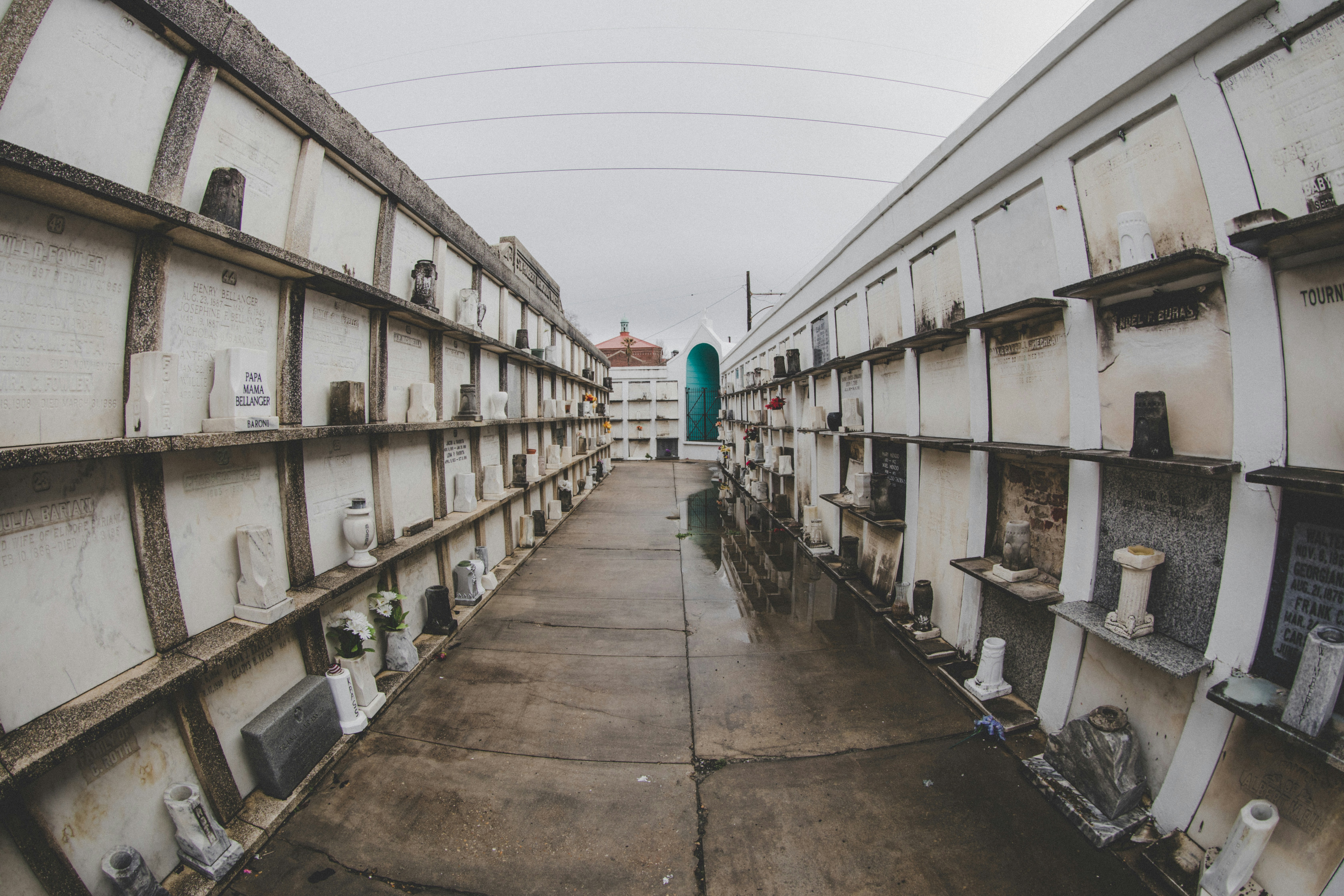 Narrow walkway flanked by mausoleums in a cemetery, leading to an ornate green entrance. The somber atmosphere reflects a place of remembrance.