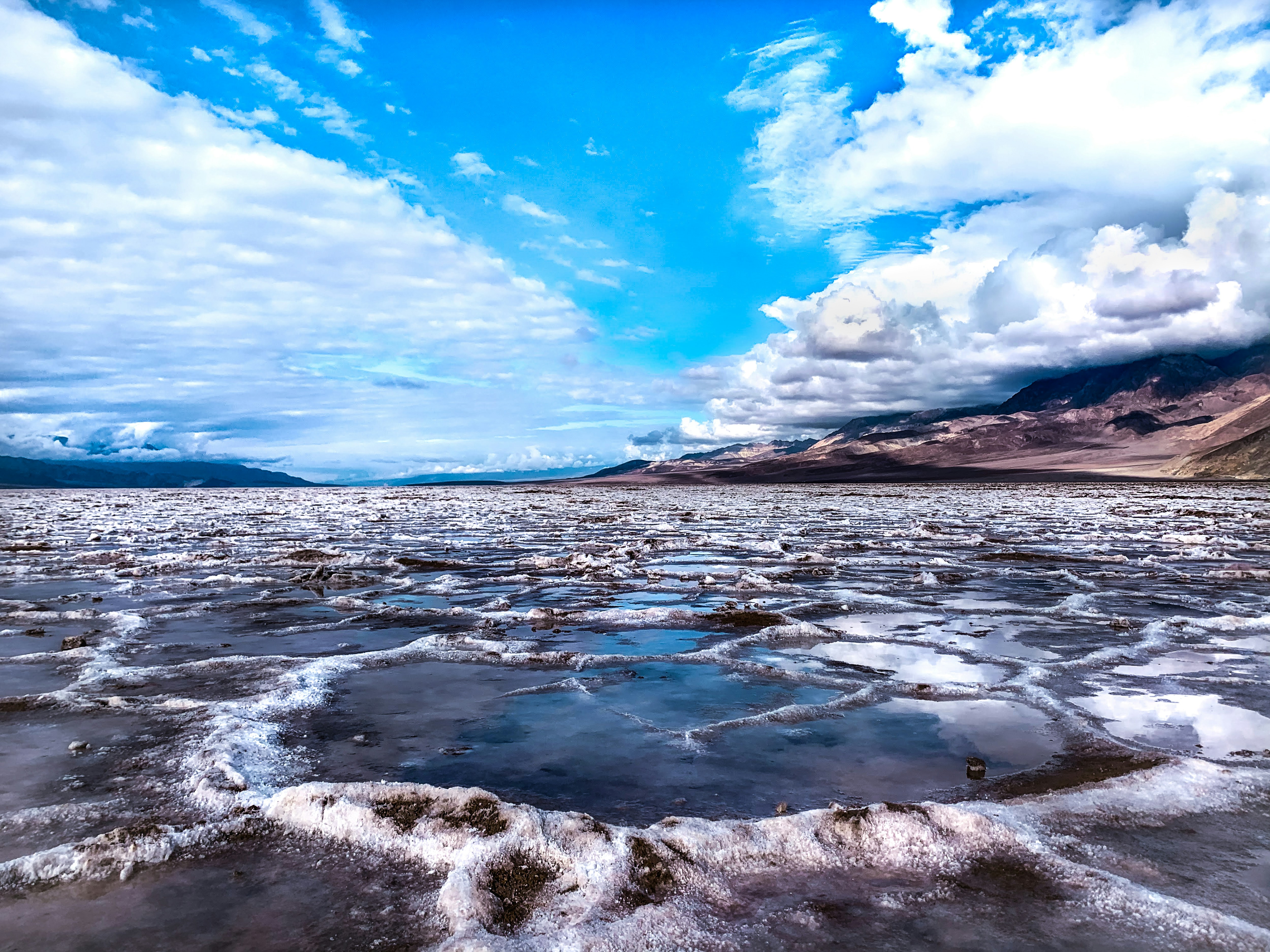 Vast salt flats stretch beneath a dynamic sky, with clouds casting shadows over the shimmering surface. The landscape showcases intricate patterns formed by natural evaporation.