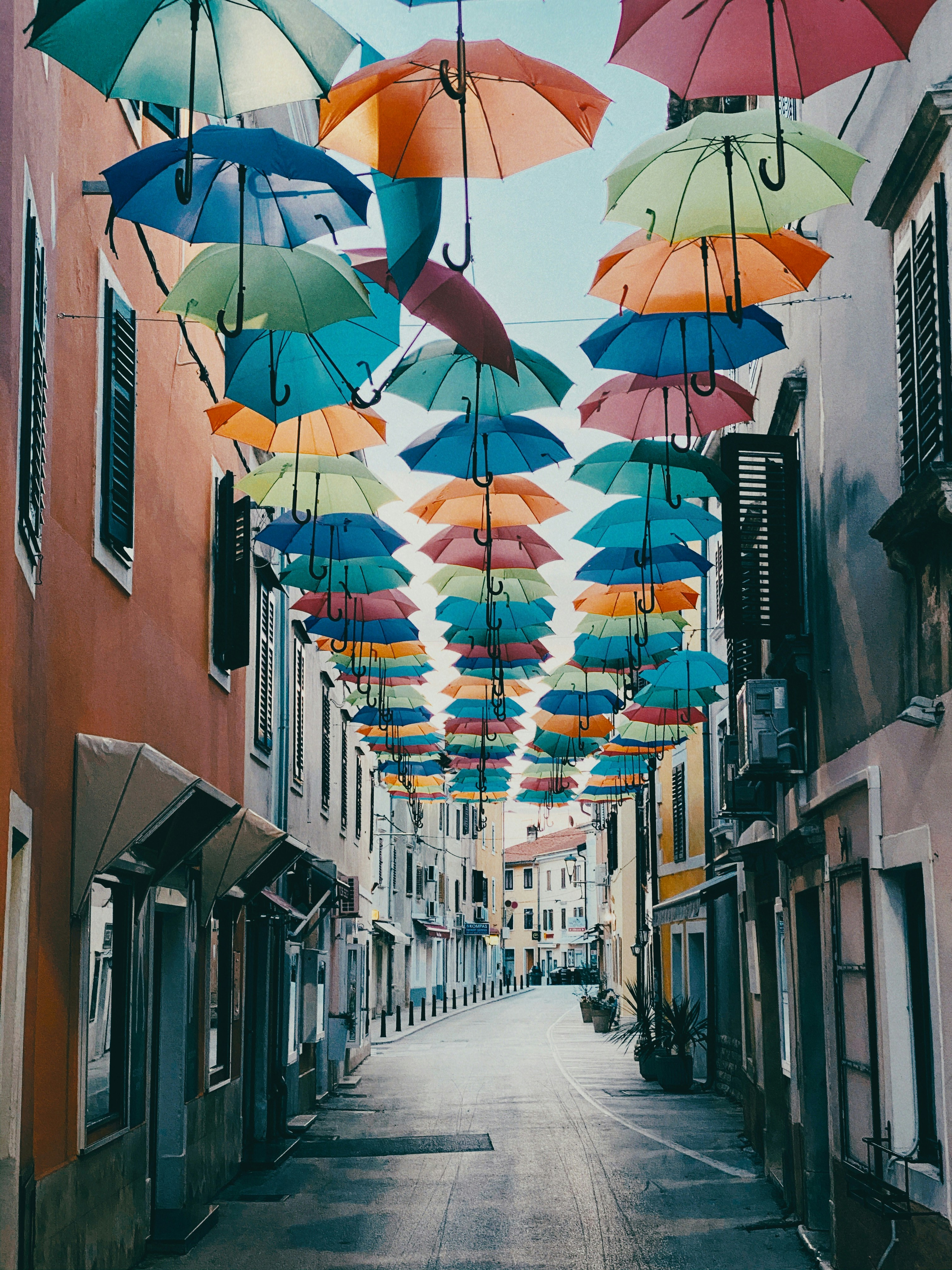 Colorful umbrellas suspended above a narrow street, creating a vibrant urban art installation. The scene showcases the charm of the surrounding architecture.