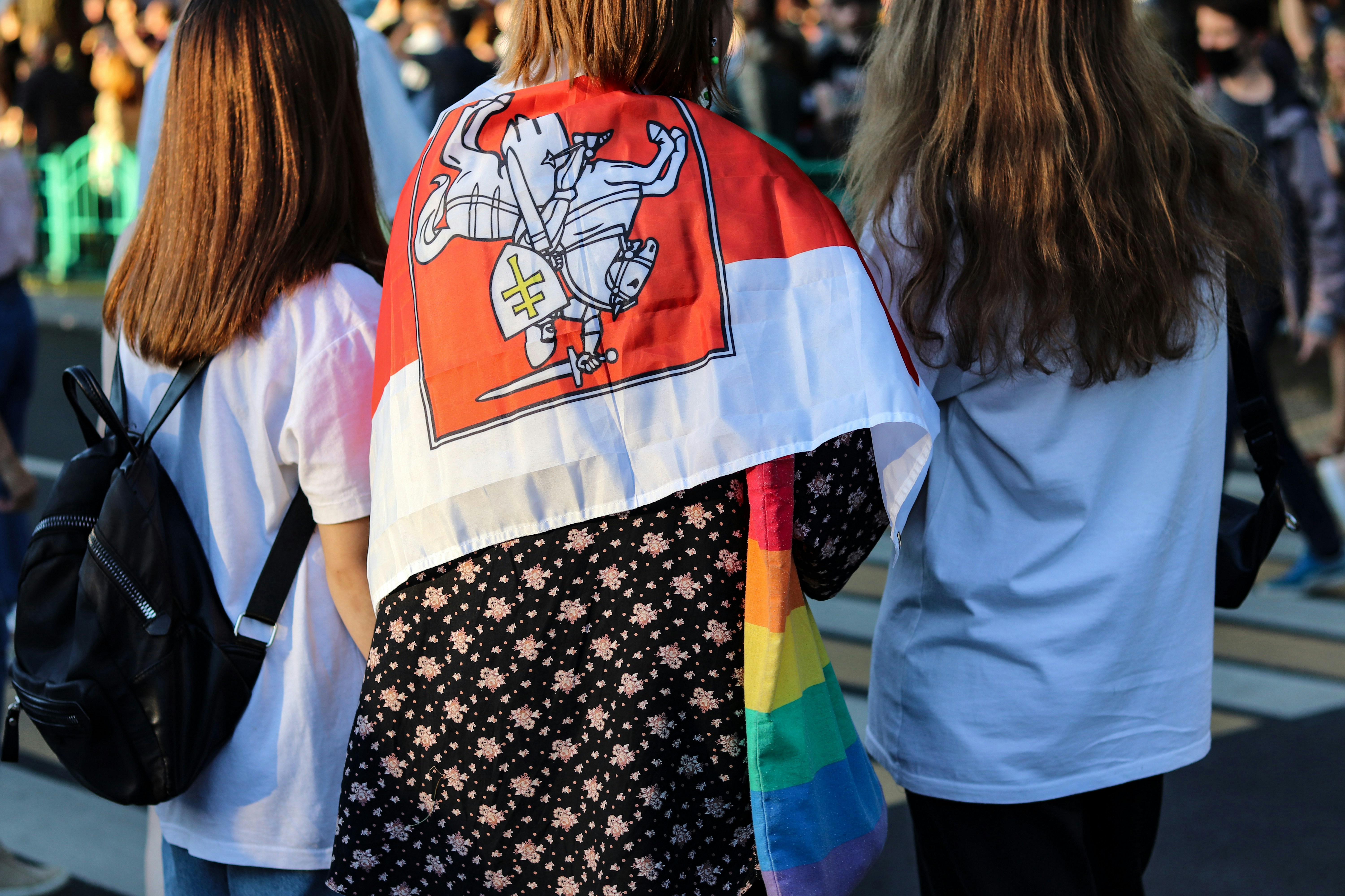 Three people standing together, one draped in a flag with a bull emblem and rainbow colors, in a crowded outdoor setting.