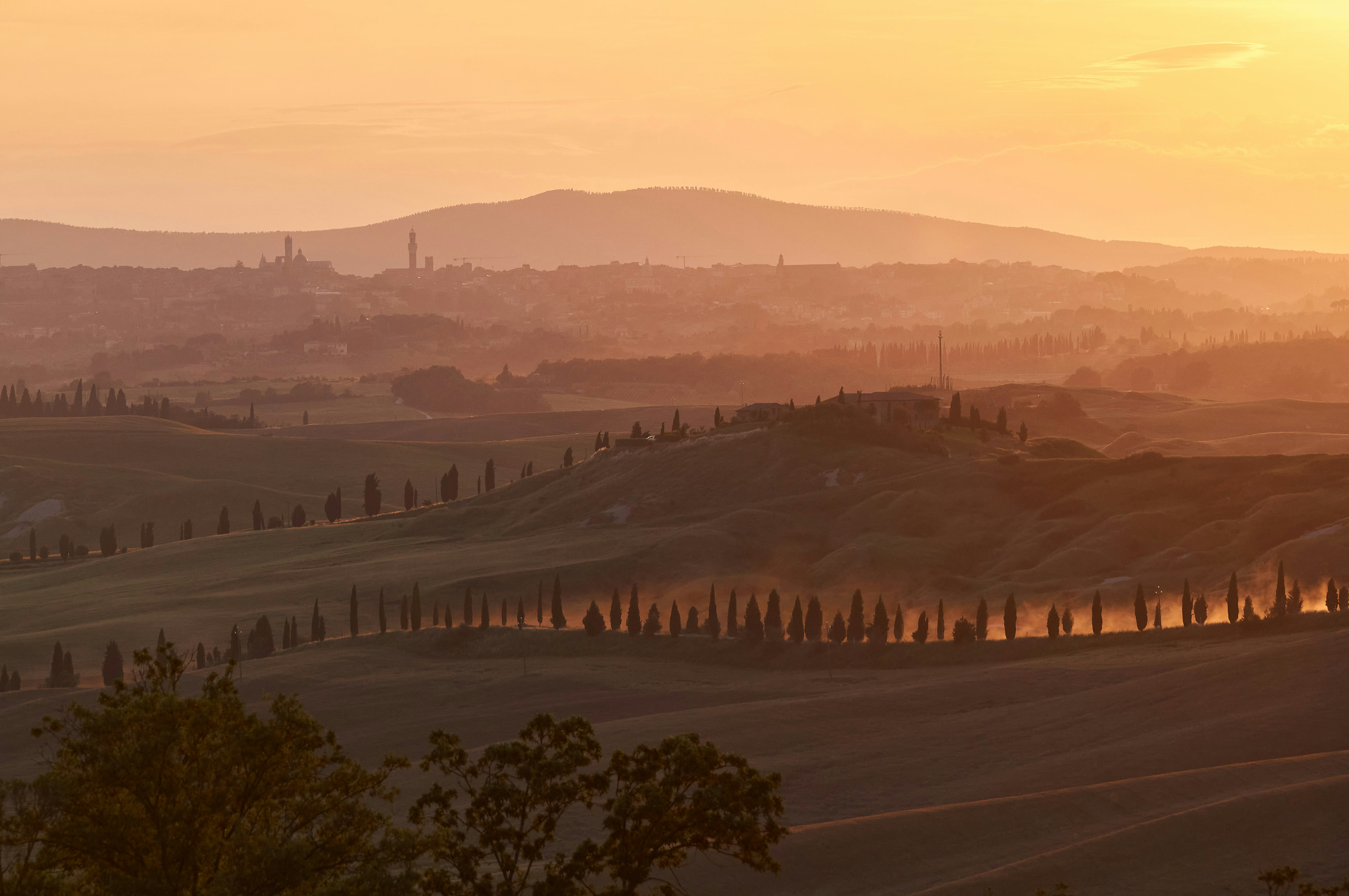 Rolling hills of Tuscany bathed in golden light, with distant silhouettes of cypress trees and a quaint village against a hazy sunset sky.
