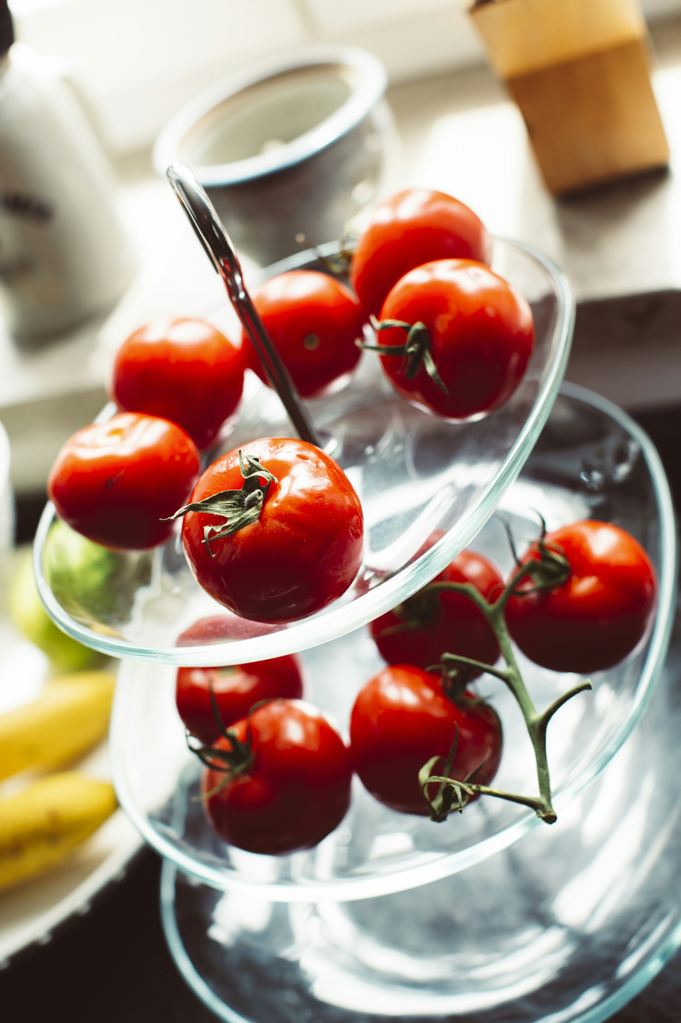 A tiered glass stand showcases freshly picked red tomatoes, glistening with freshness, surrounded by hints of other fruits. The scene captures the essence of a homey kitchen atmosphere.