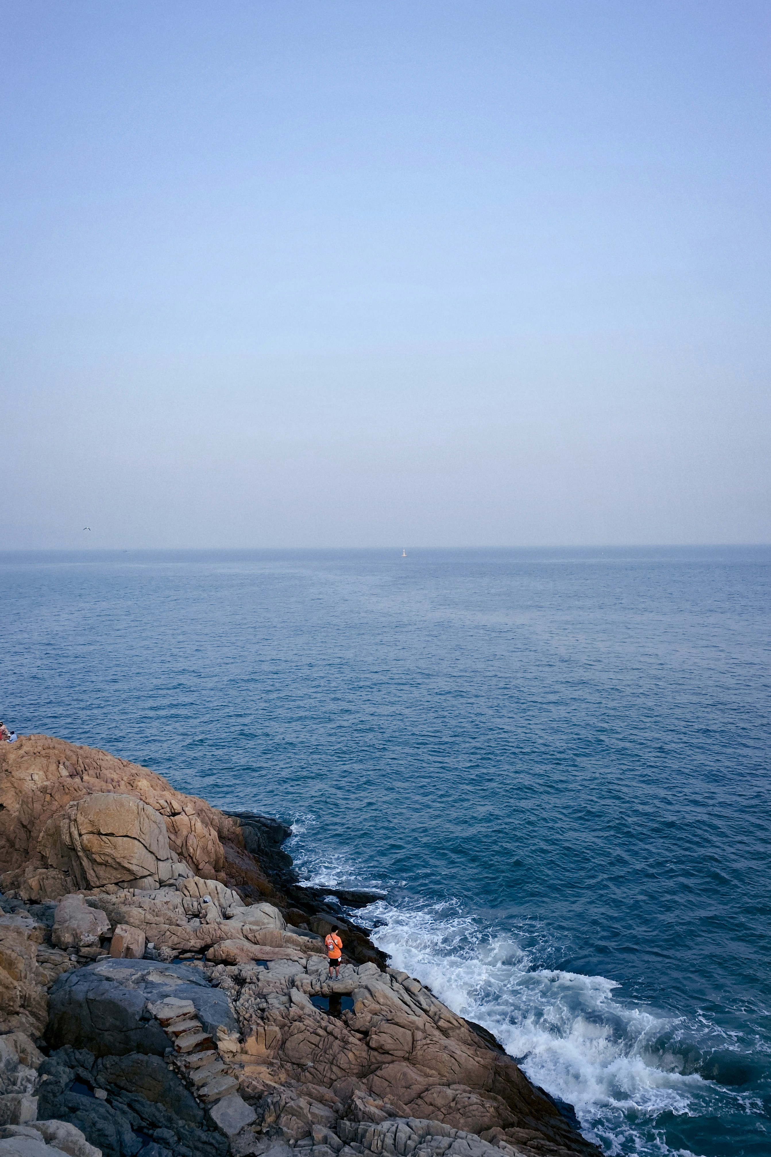 A lone figure stands on rocky shores, gazing at the tranquil sea under a soft blue sky. Waves gently lap against the stones.