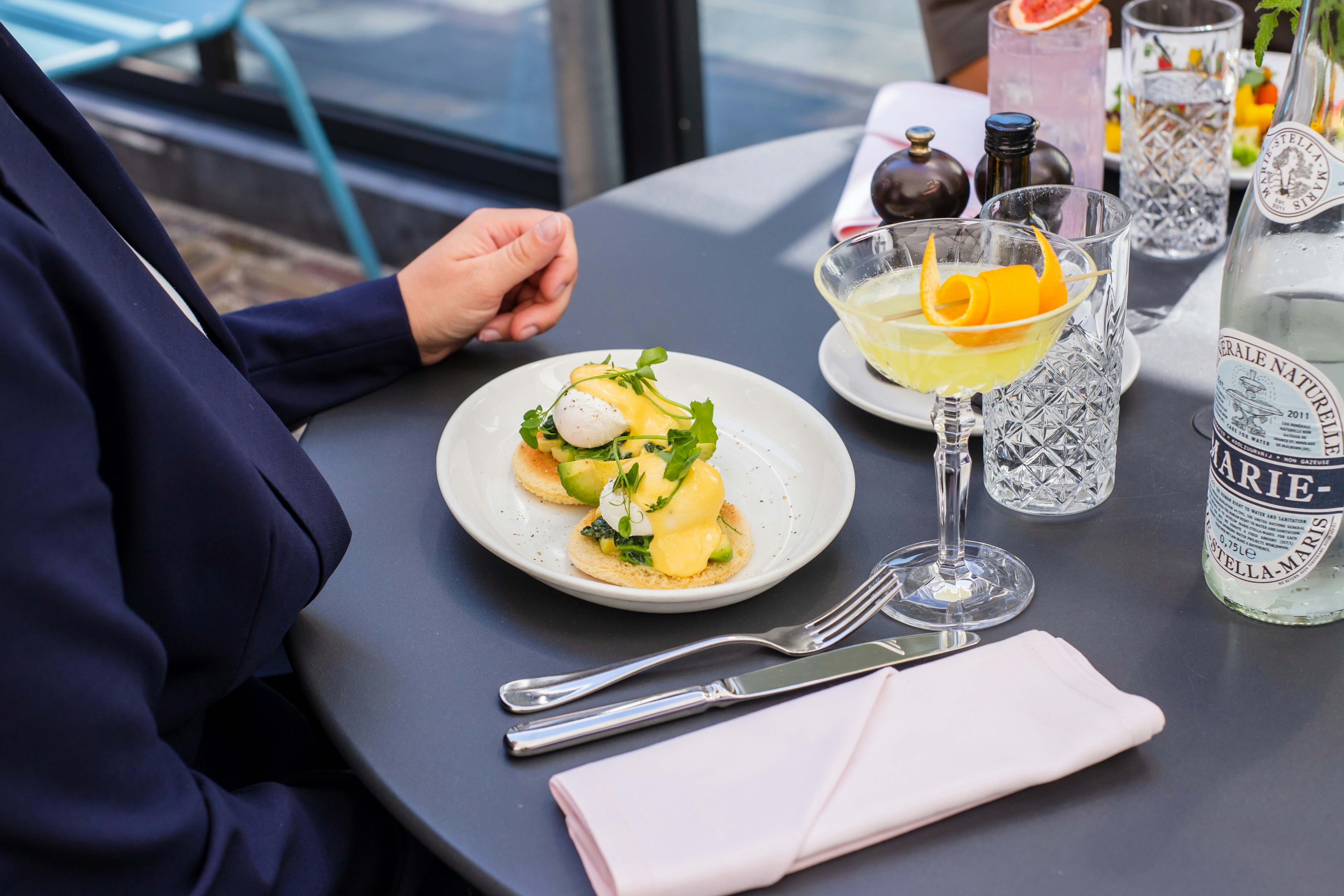 A table set with poached eggs on toast and a cocktail, next to a bottle and glassware.