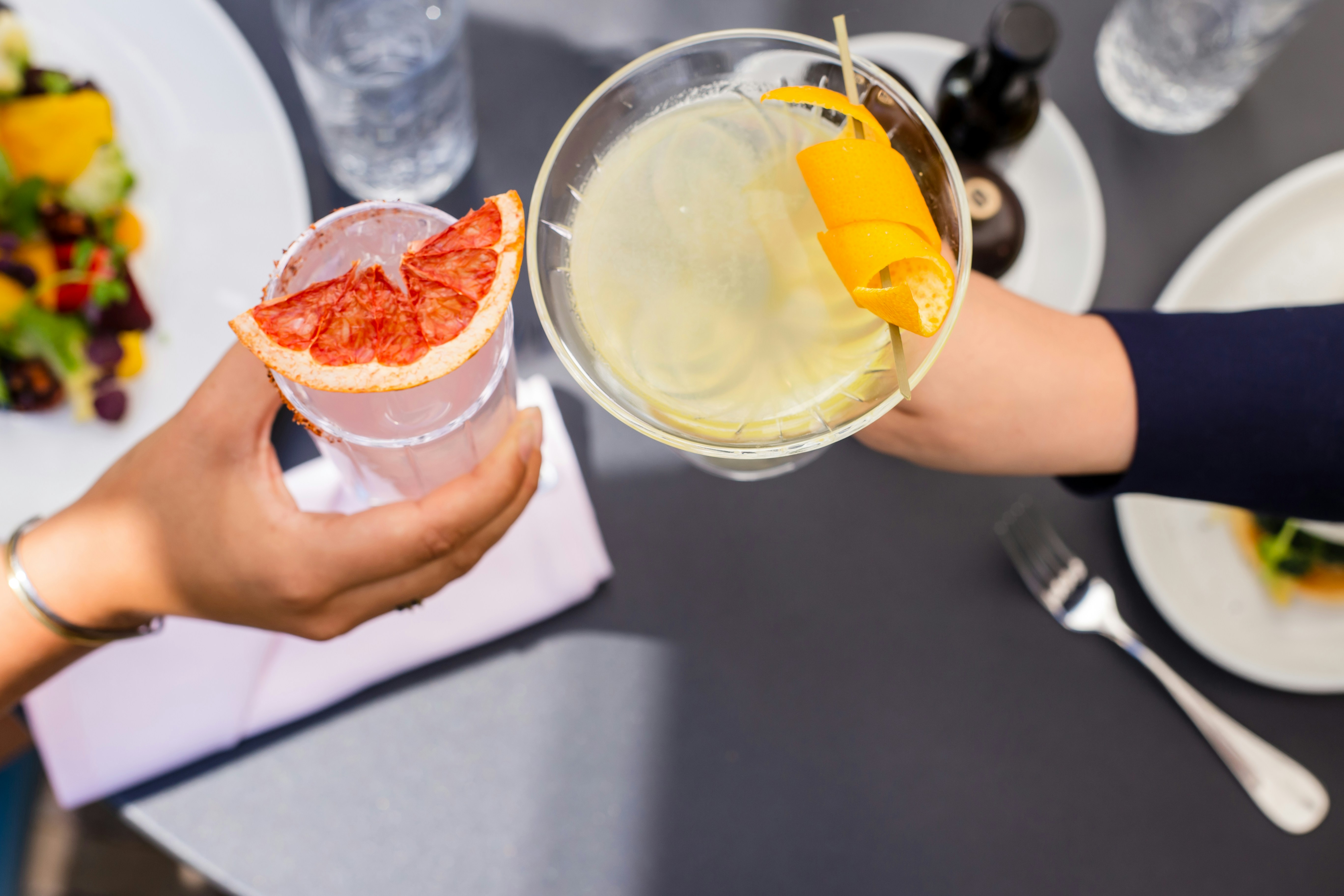 Two hands toasting with grapefruit and lemon garnished cocktails over a table setting.