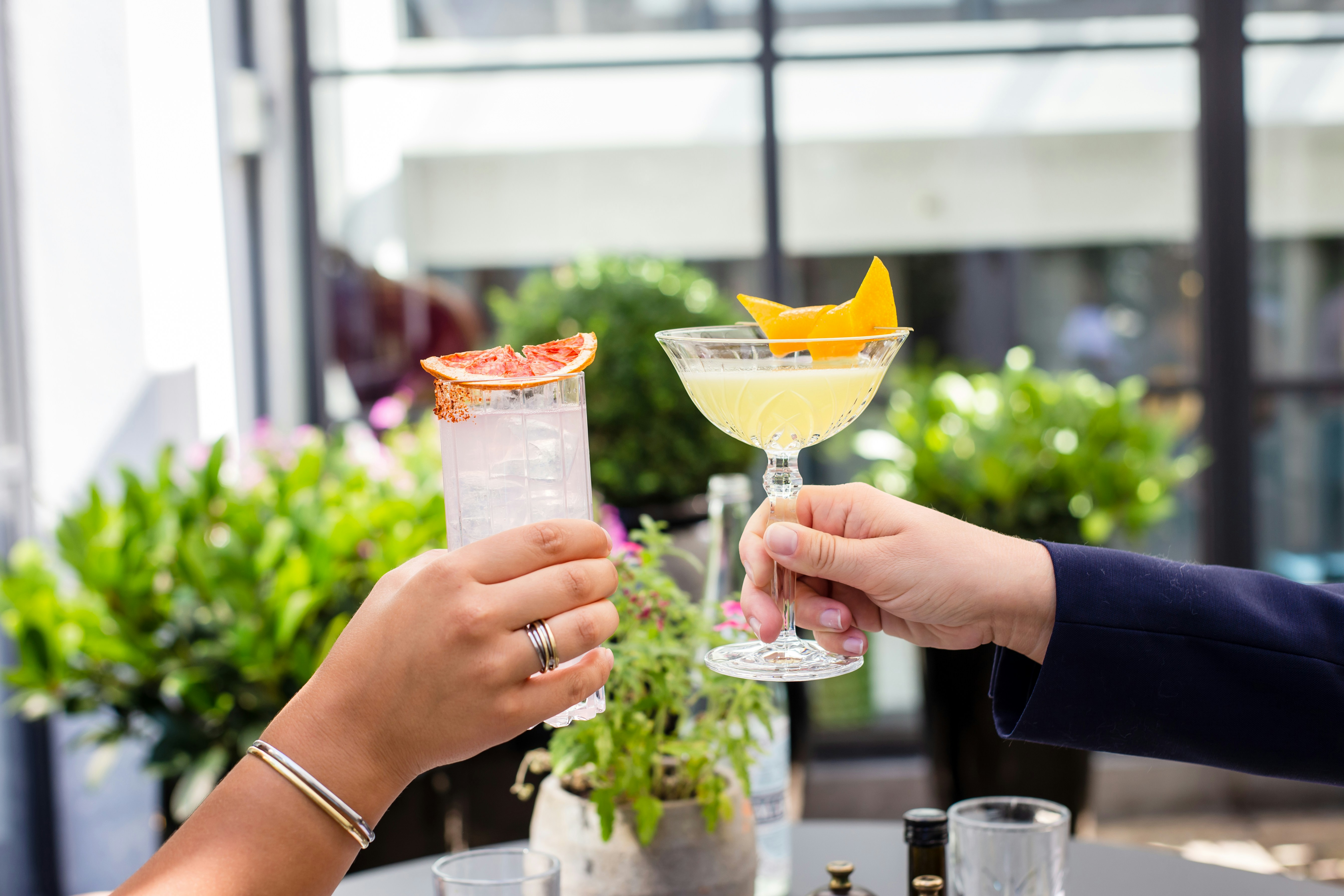 Two hands clink cocktails against a backdrop of lush greenery and modern architecture.