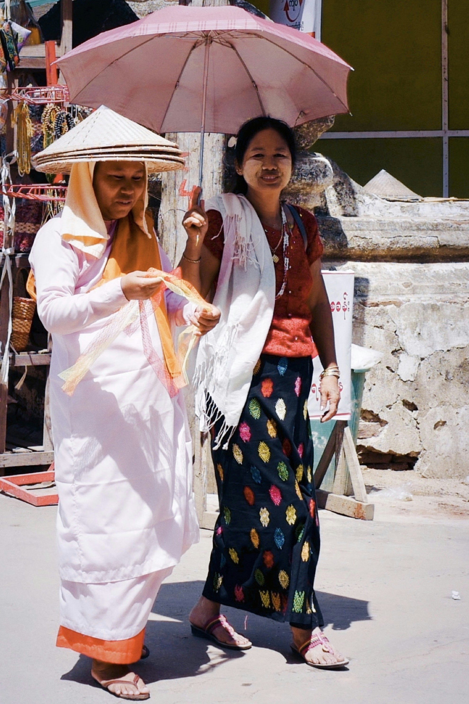 Two women stroll through a vibrant market, one holding a colorful fabric while the other shields herself with an umbrella. Their traditional attire reflects local craftsmanship.