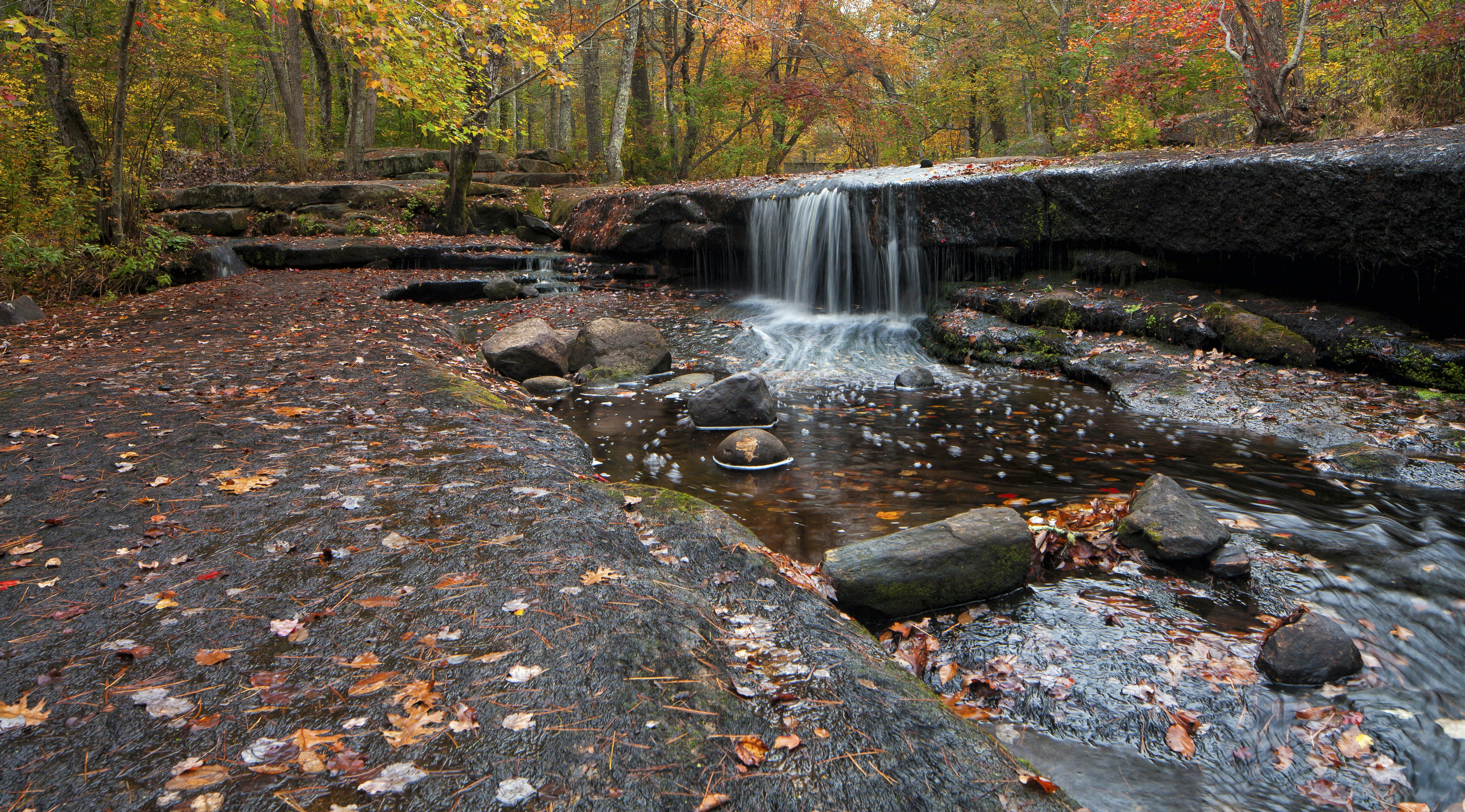 Waterfall gently flowing over flat rocks surrounded by colorful autumn foliage.