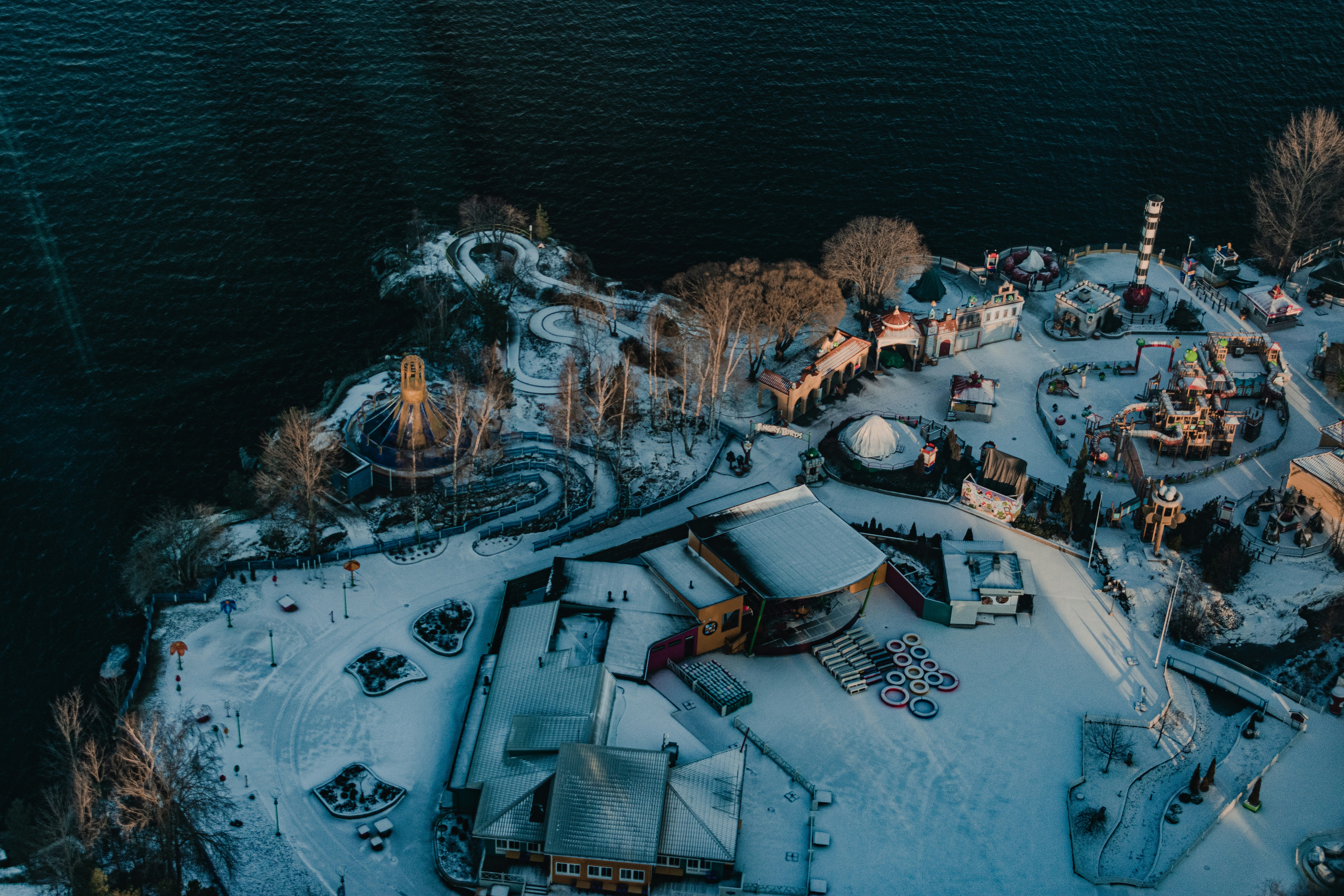 Aerial view of a snow-covered amusement park with rides and attractions, framed by a serene body of water. The scene captures the playful essence of winter leisure.