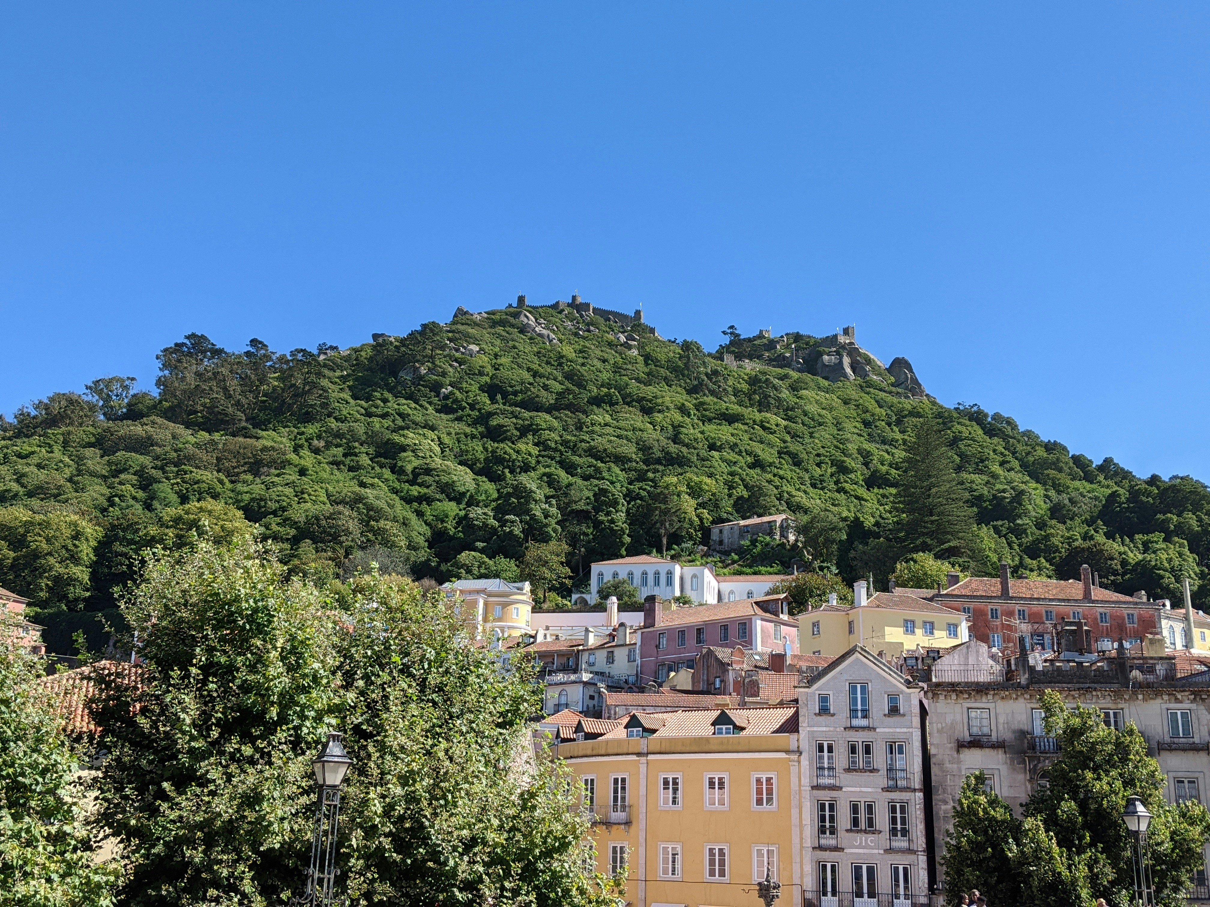 Vibrant hillside town with pastel buildings nestled against a lush green hill under a clear blue sky.
