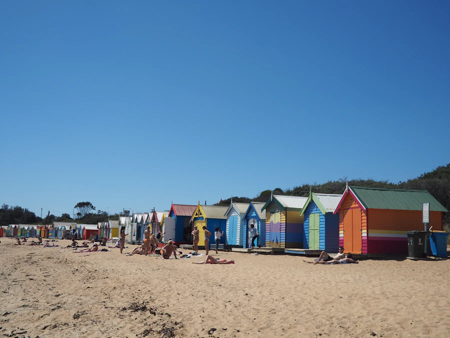 A row of brightly painted beach huts lines a sandy shore under a clear blue sky, with people relaxing nearby. The scene feels cheerful and summery.