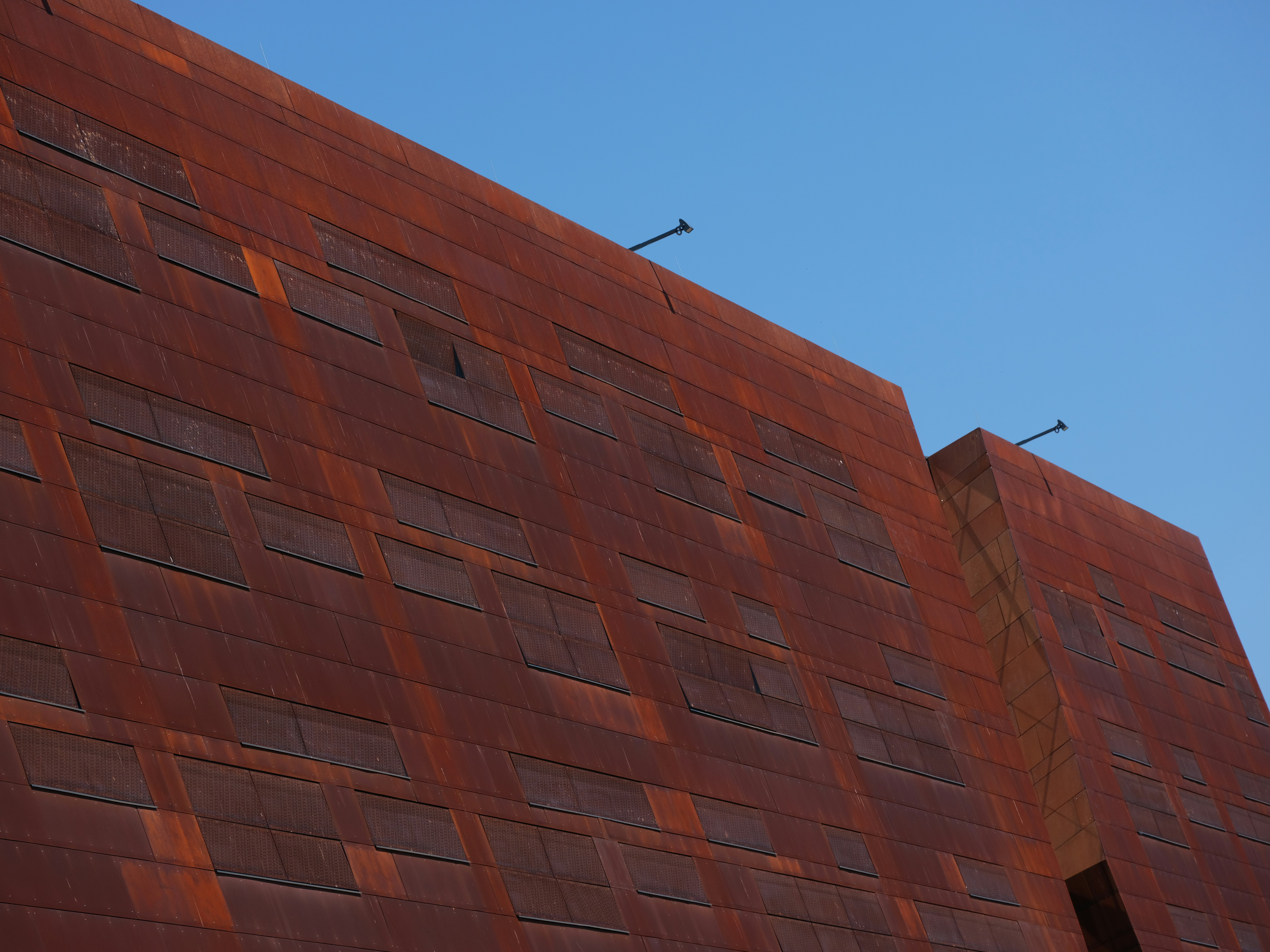Abstract view of a weathered, rust-colored building facade against a clear blue sky. The geometric patterns create a striking visual contrast.