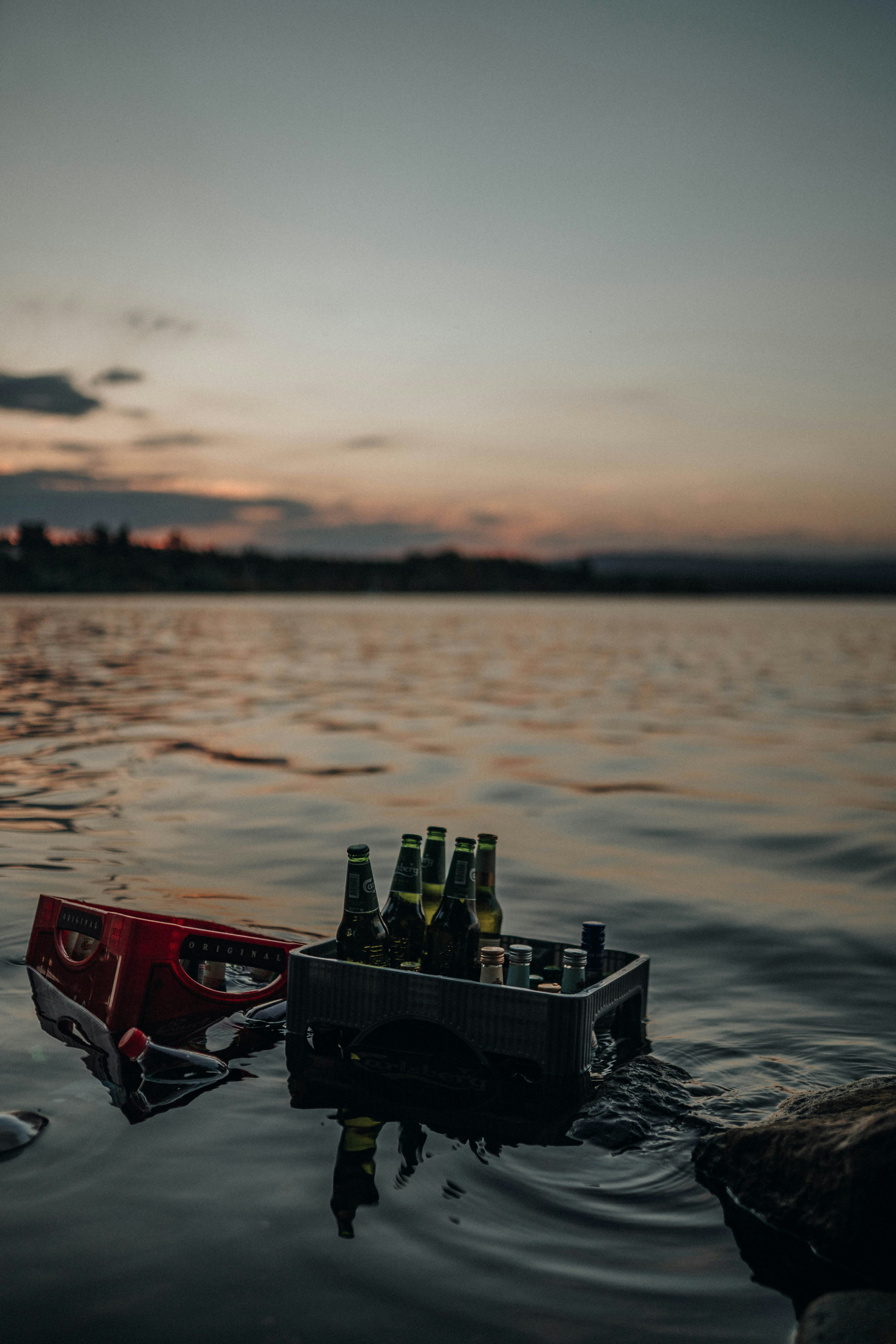 Two crates filled with assorted bottles float gently on a tranquil lake at dusk, reflecting the soft hues of the evening sky.