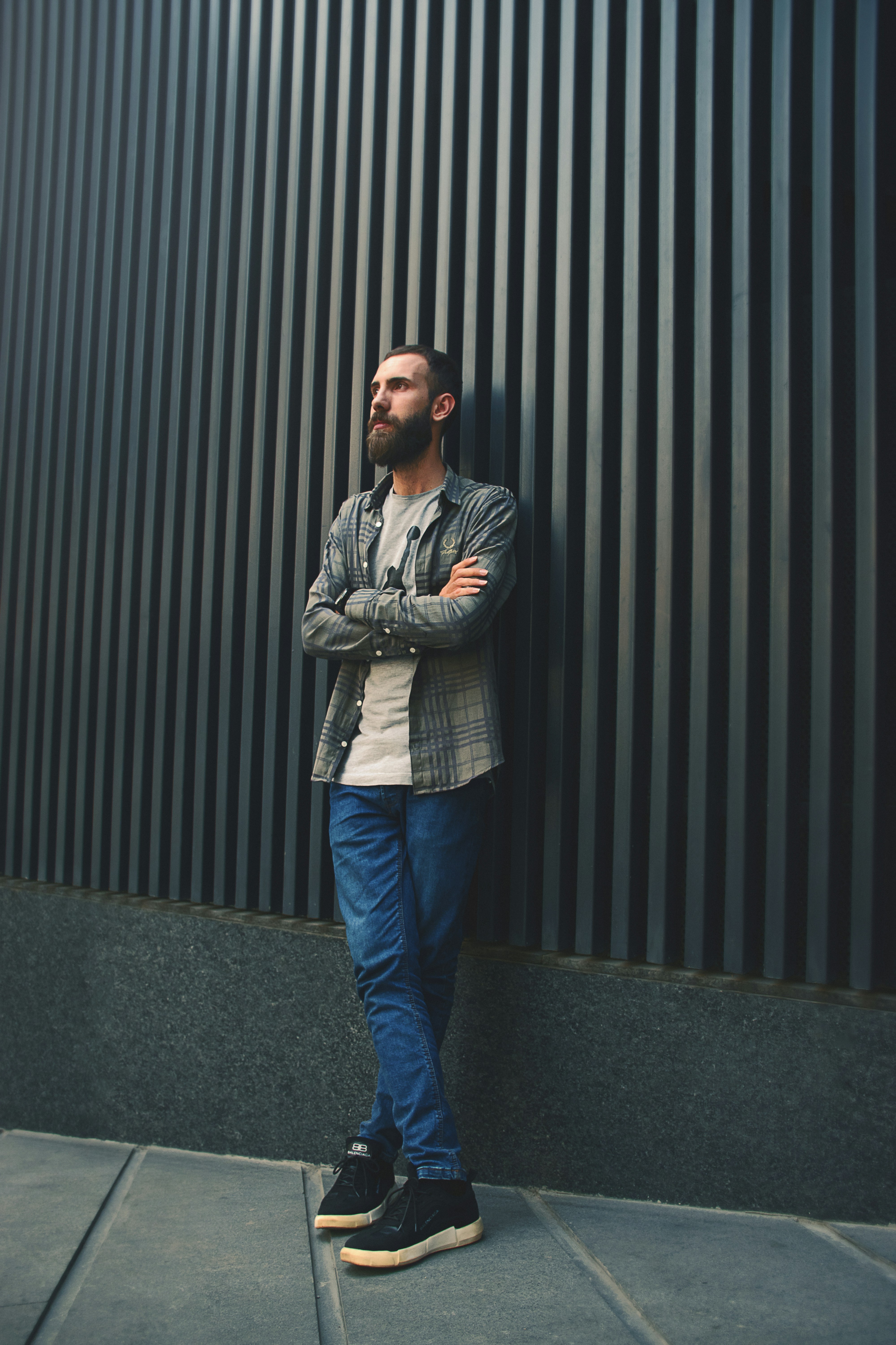 A man with a beard stands confidently against a vertical striped wall, arms crossed, exuding a relaxed yet thoughtful demeanor.