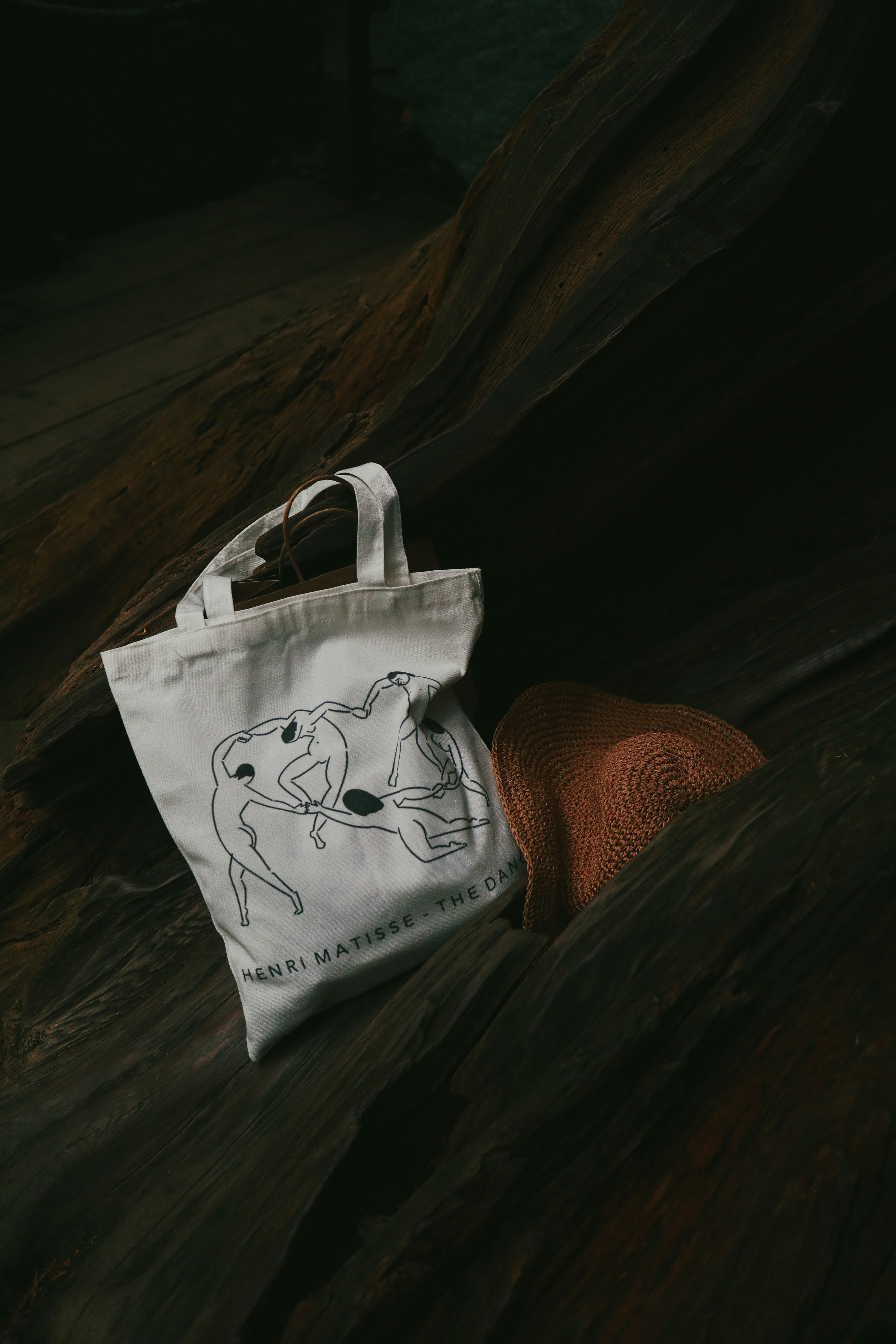 Canvas tote bag featuring a line drawing inspired by Henri Matisse, resting on a textured wooden surface, accompanied by a woven hat.
