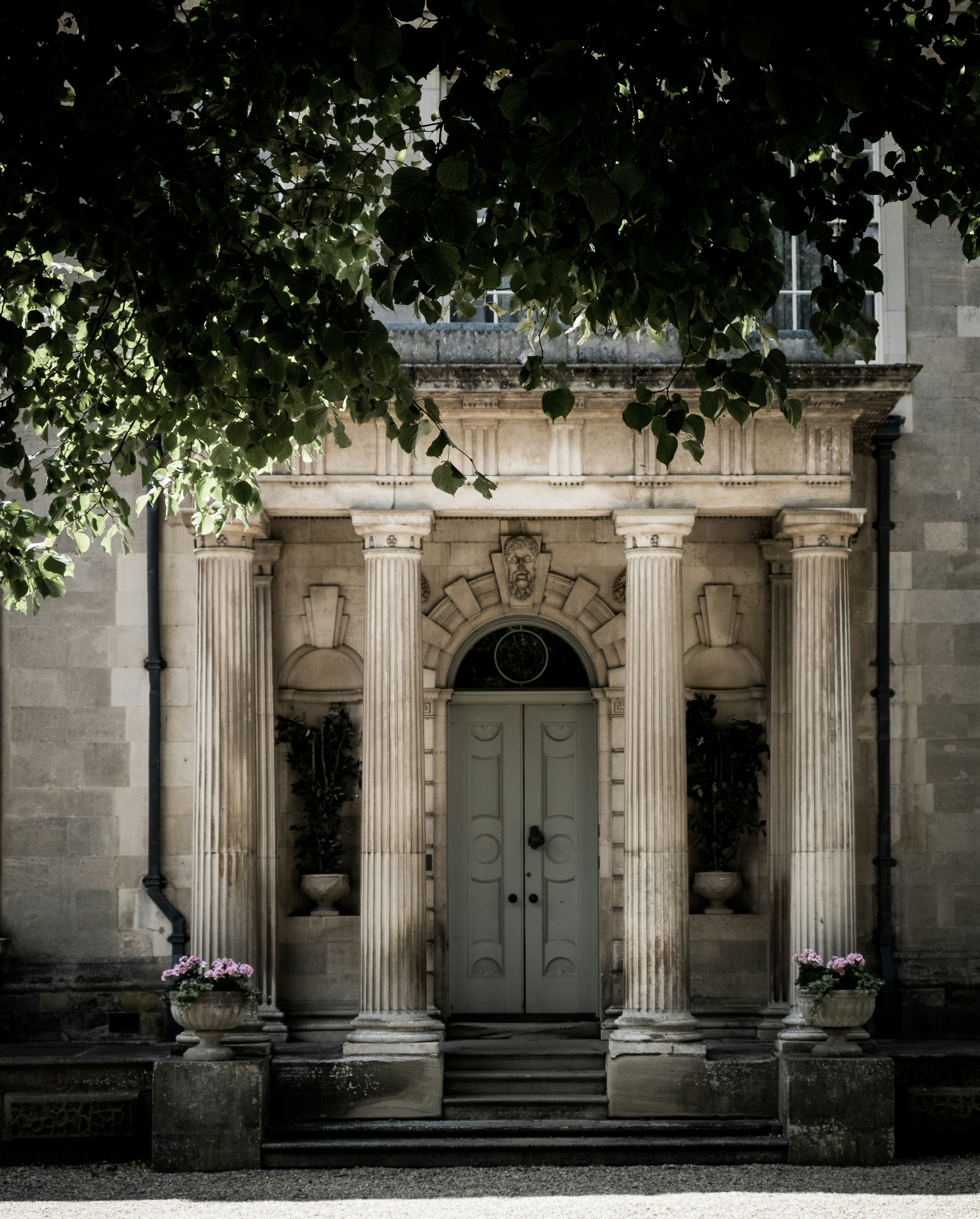 Classical stone entrance framed by lush tree branches casting dappled shadows.