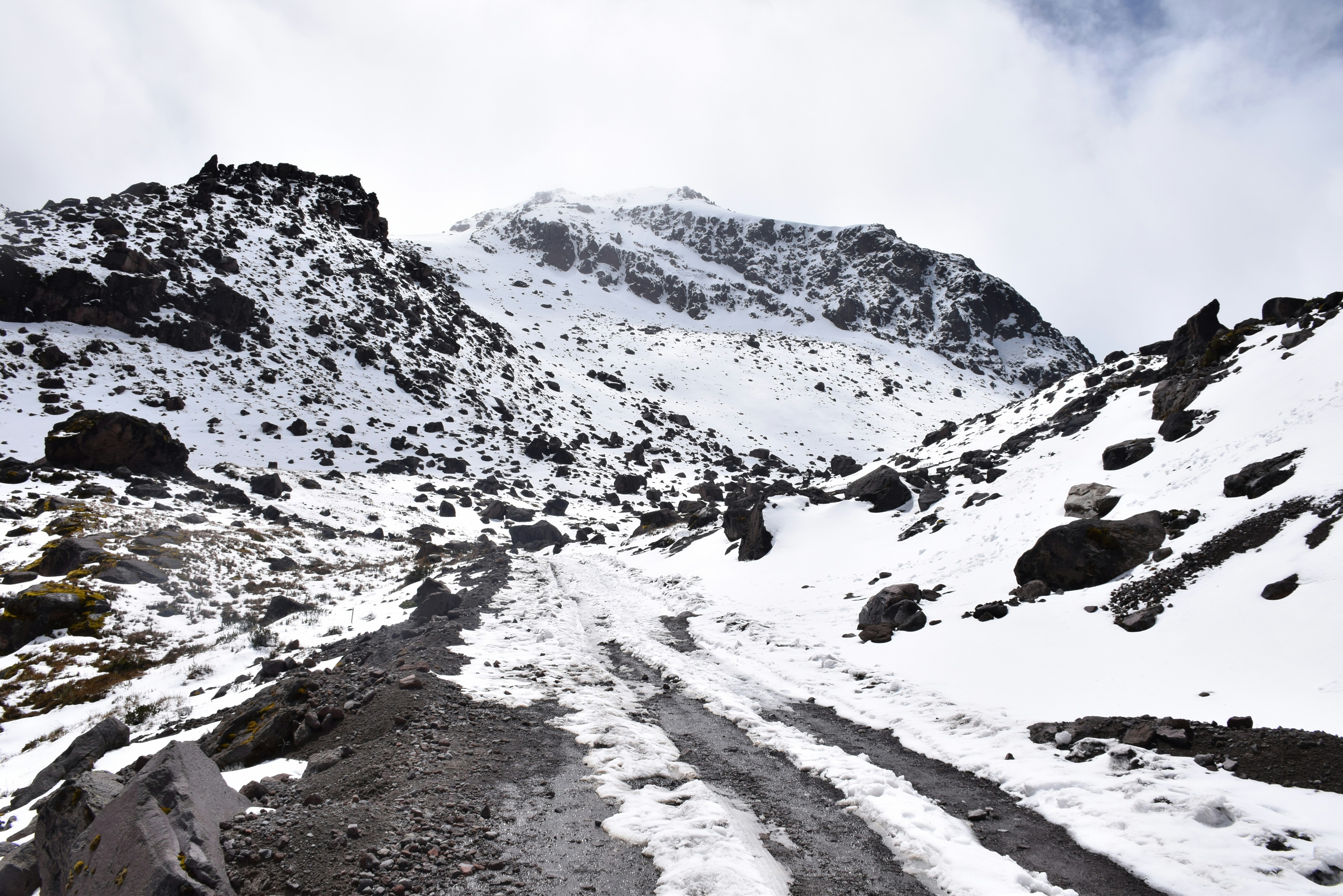 Snow-laden path winding through rocky terrain towards Cayambe Volcano under a cloudy sky.