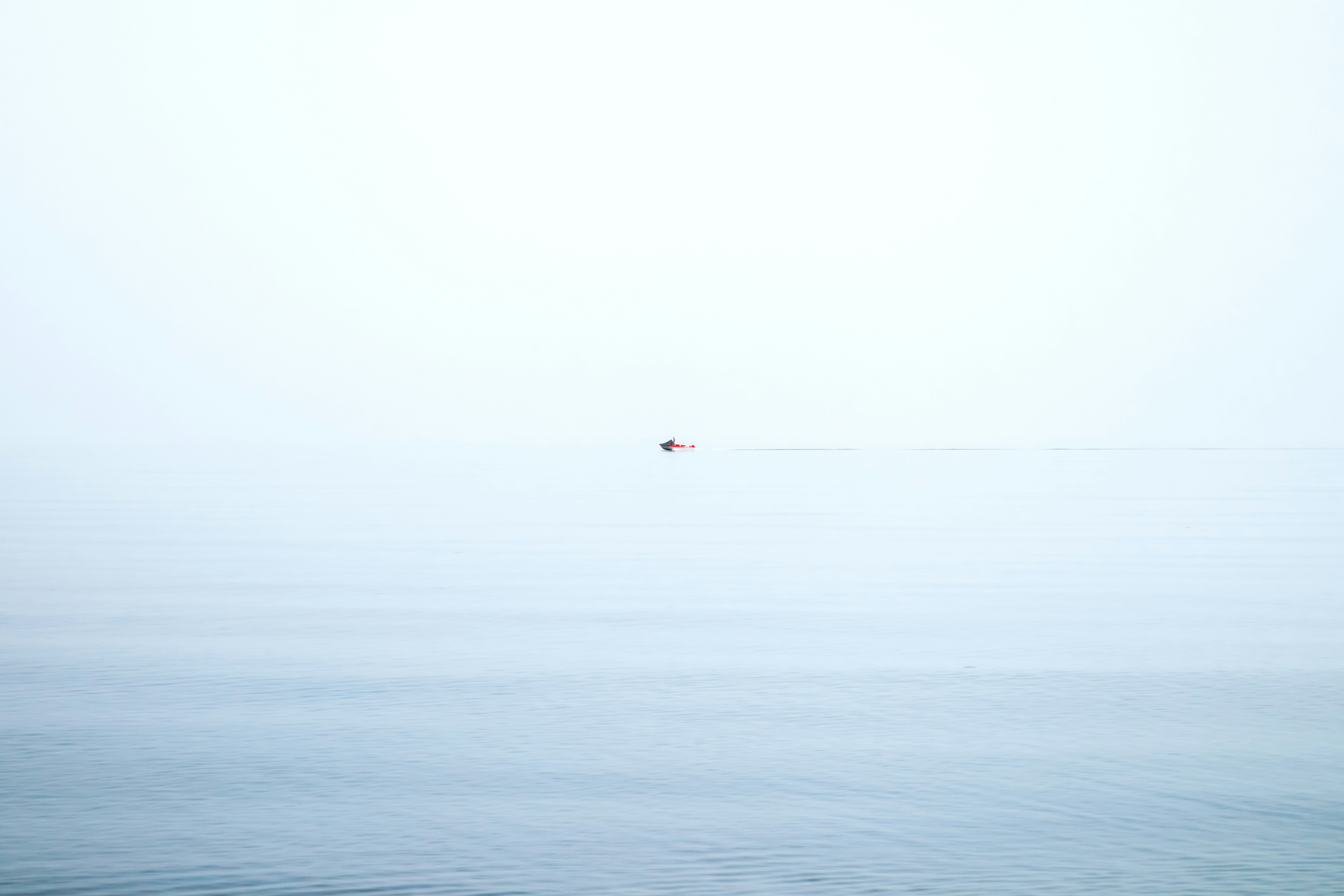 A lone red boat drifts across a tranquil, misty sea, creating a serene atmosphere. The soft color palette enhances the feeling of calm.