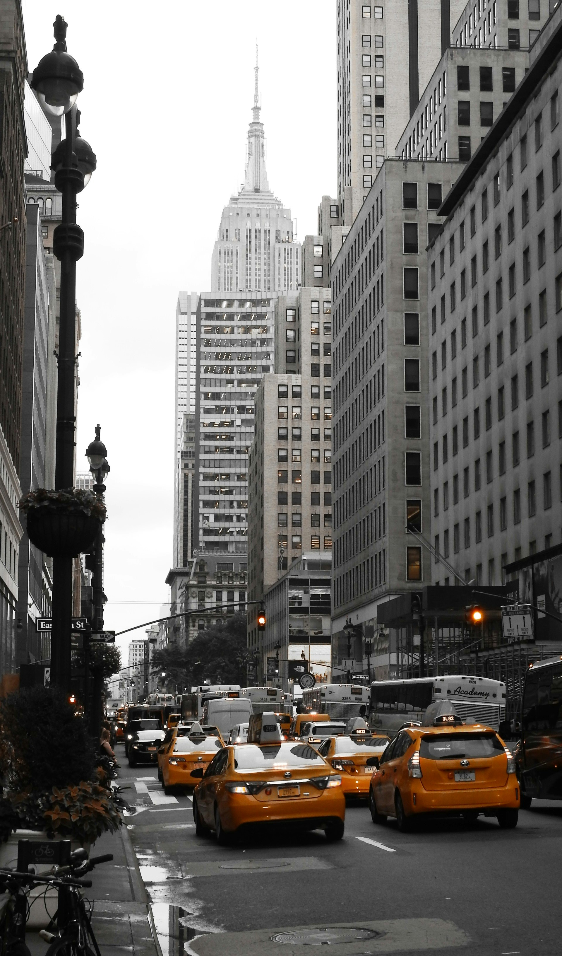 Yellow taxis navigate a bustling city street, framed by towering skyscrapers and the iconic Empire State Building. The scene captures the vibrant energy of urban life.