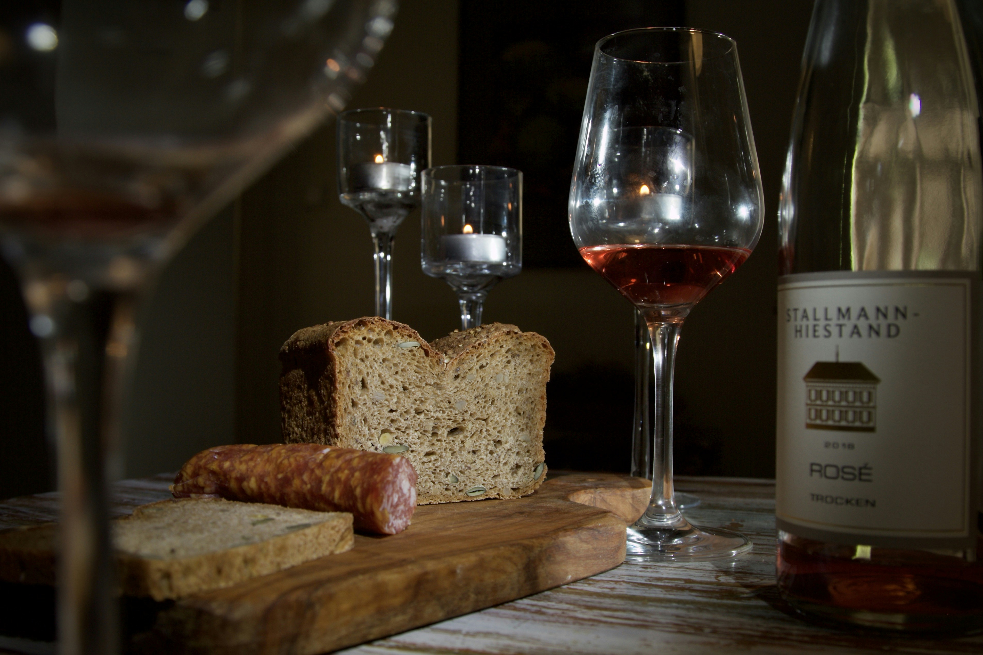 Slices of bread and sausage on a wooden board with wine glasses and a bottle in the background.