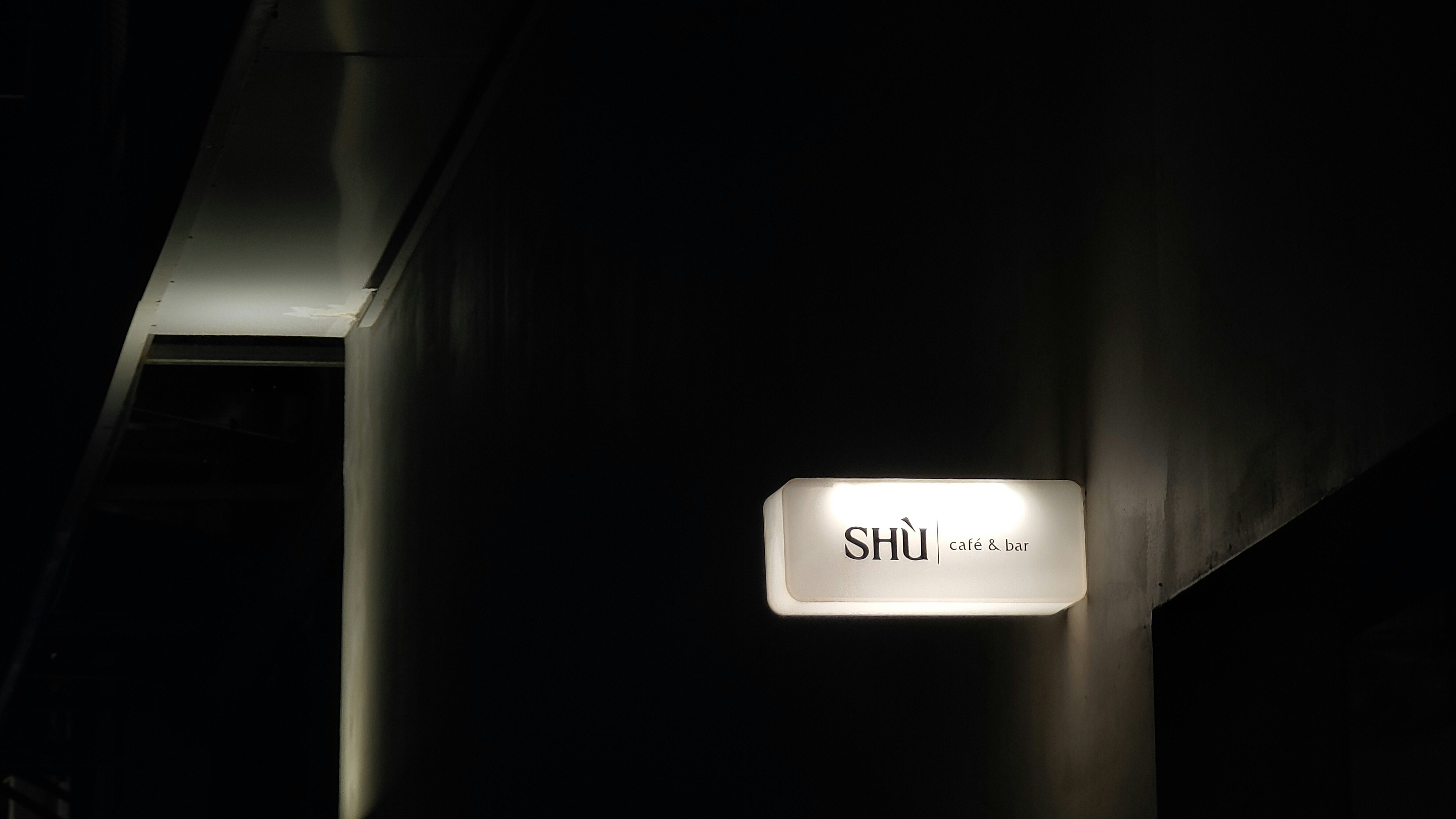 Dim hallway with a softly illuminated SHU sign on the wall. The sign functions as a subtle logo amid moody architectural textures.