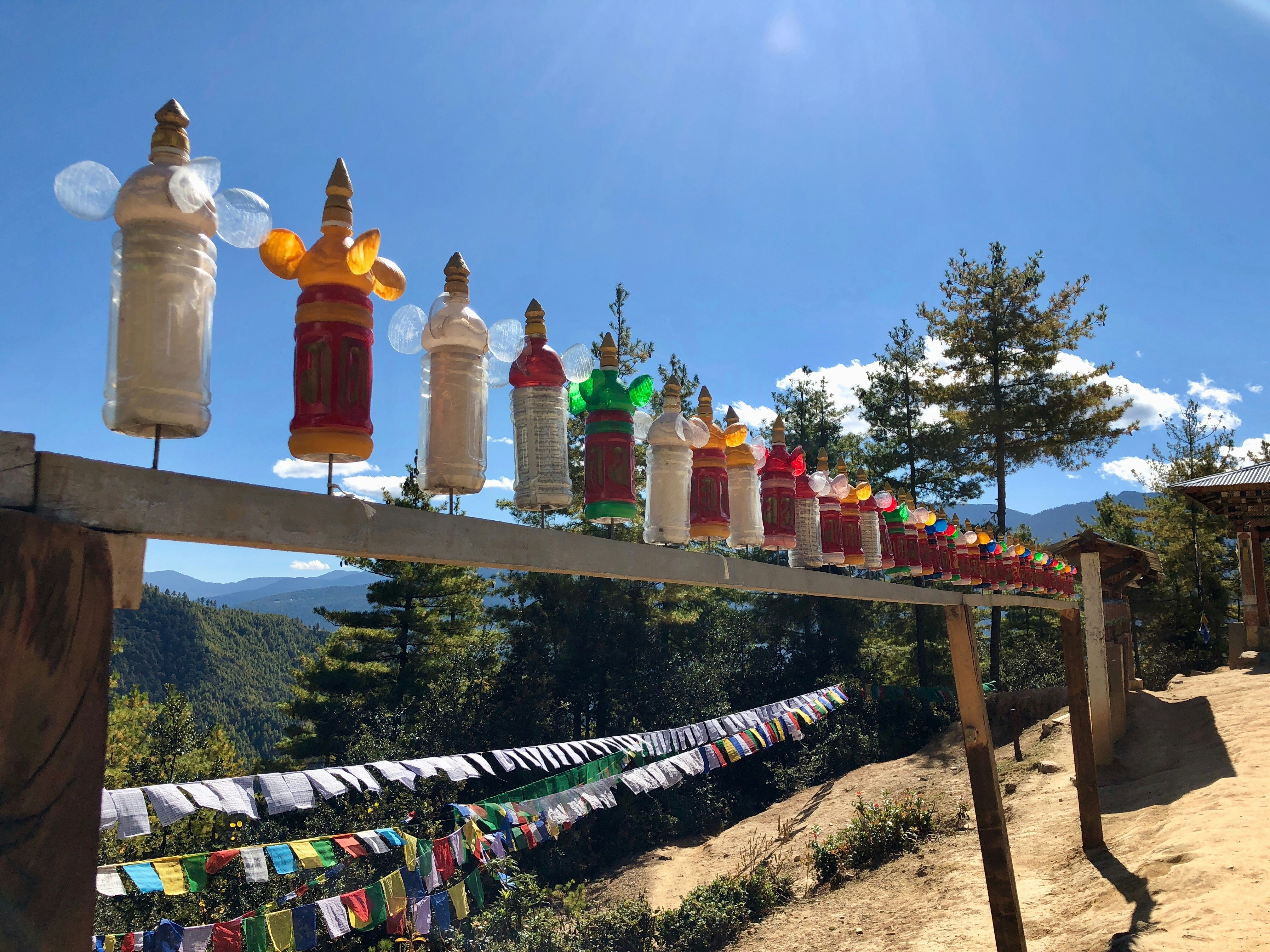Colorful prayer flags and decorative bottles line a wooden railing, set against a backdrop of serene mountains and clear skies.