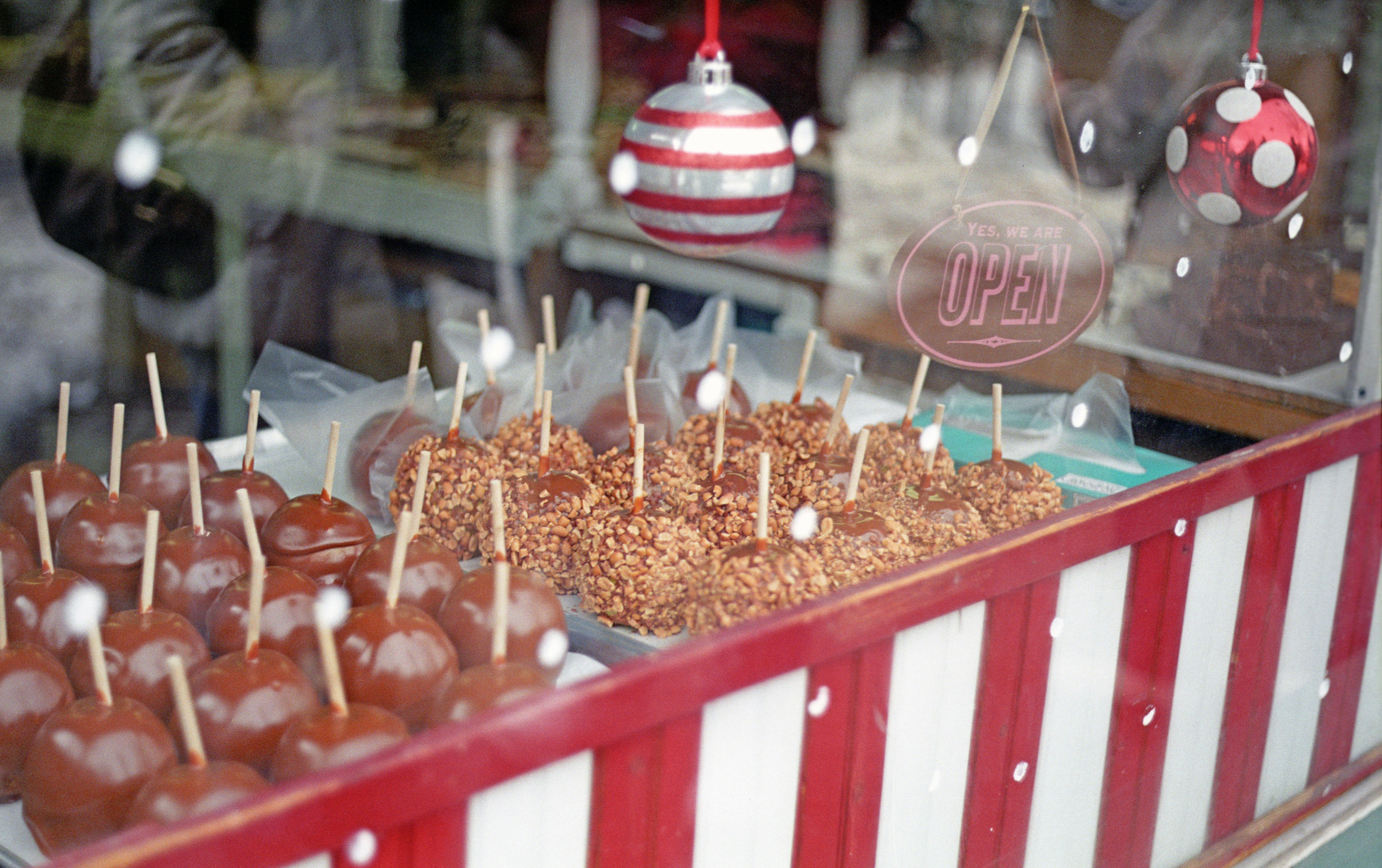 A display of caramel and nut-covered apples in a festive shop window, adorned with red and white ornaments. The scene radiates a warm, inviting atmosphere.