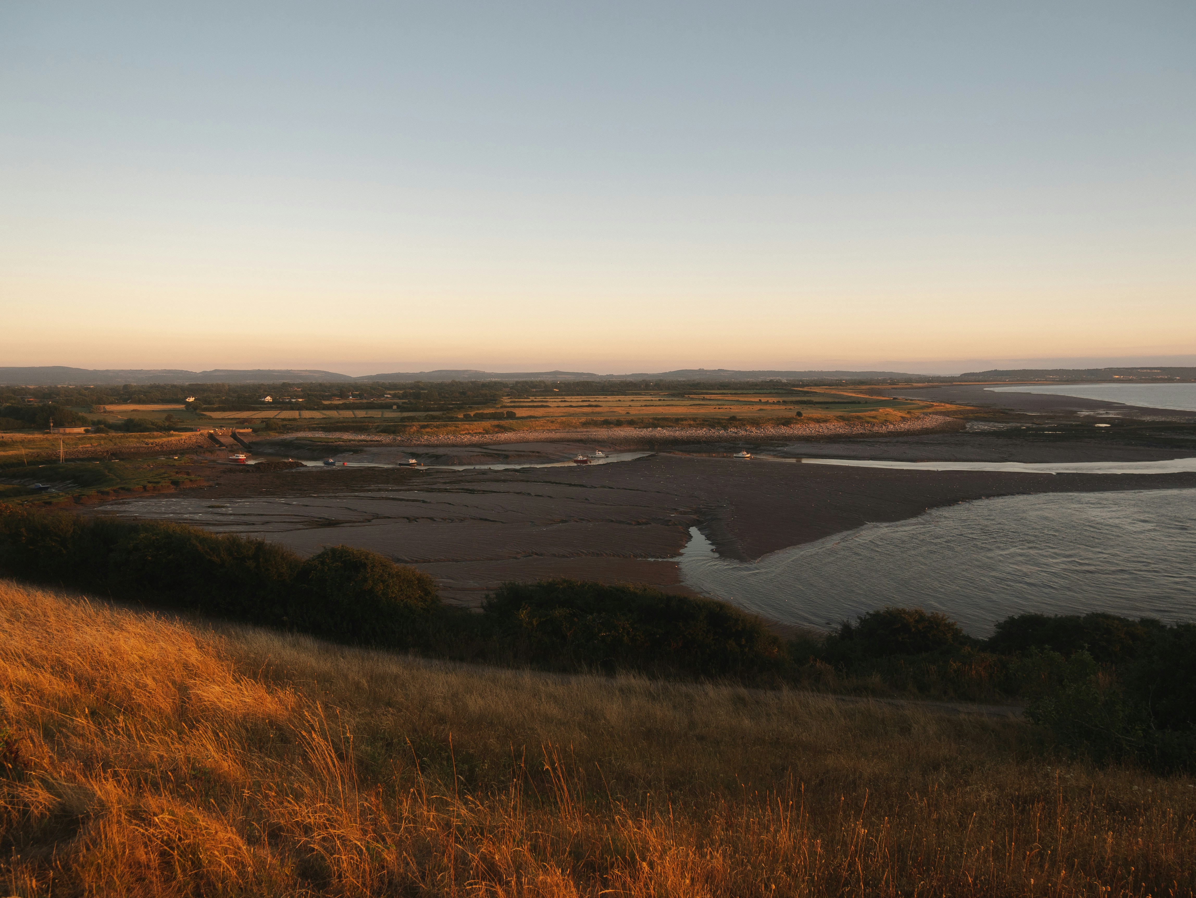 Sunset casts a warm glow over a coastal estuary with grassy hills in the foreground and calm waters stretching to the horizon.