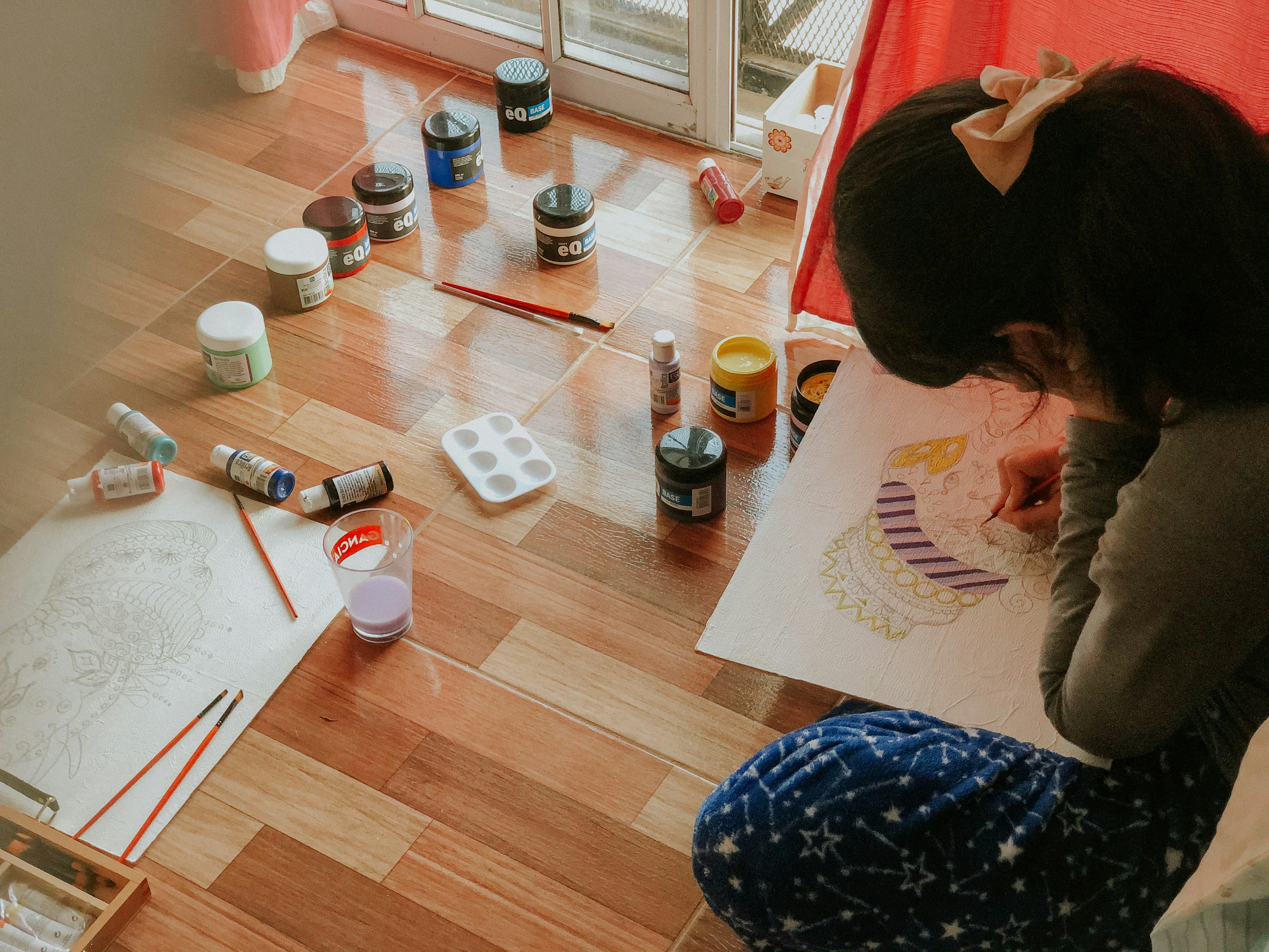Woman in pajamas painting on the floor surrounded by art supplies in natural light.