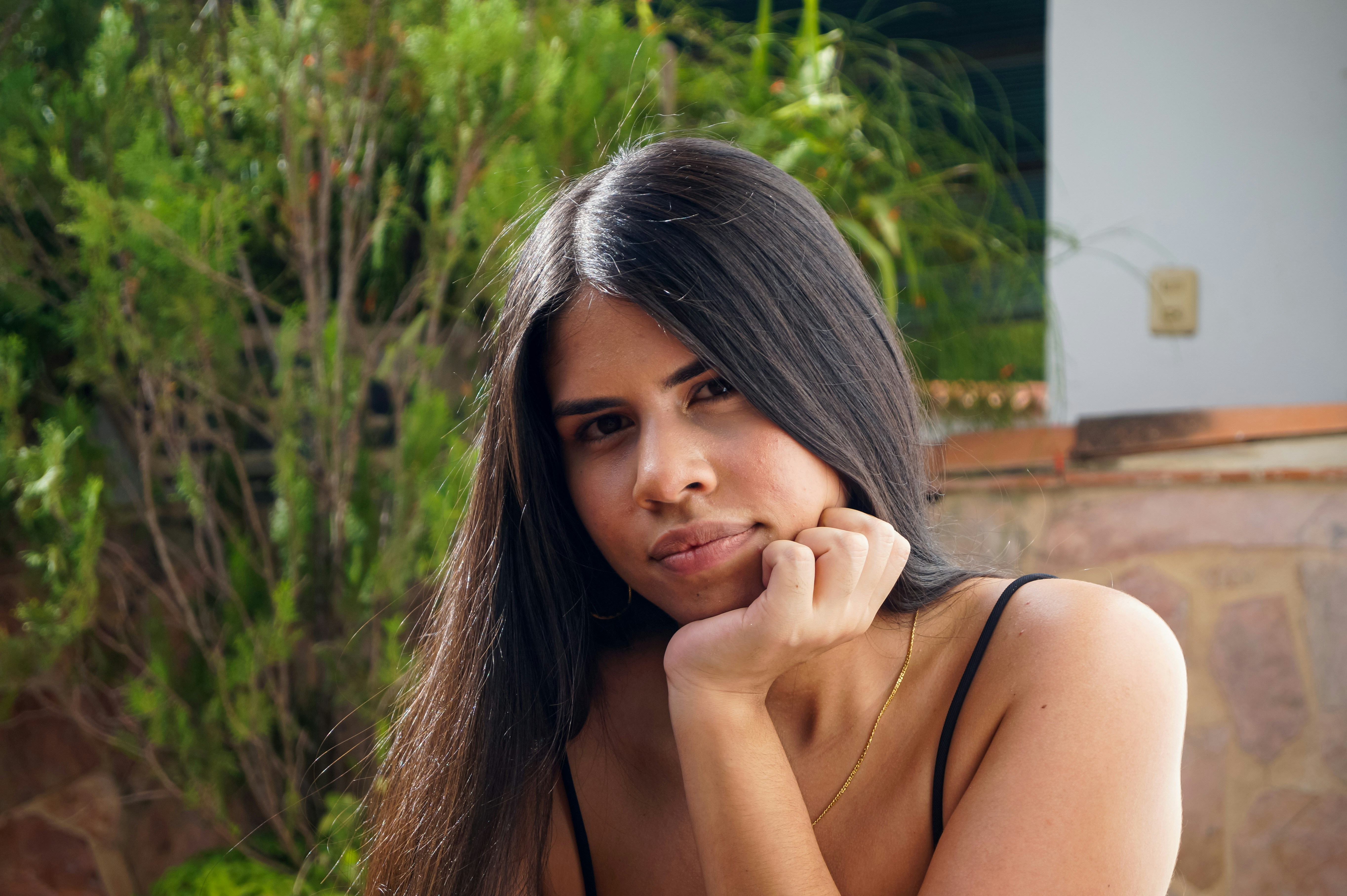 Woman with long dark hair resting her chin on her hand, surrounded by lush greenery.