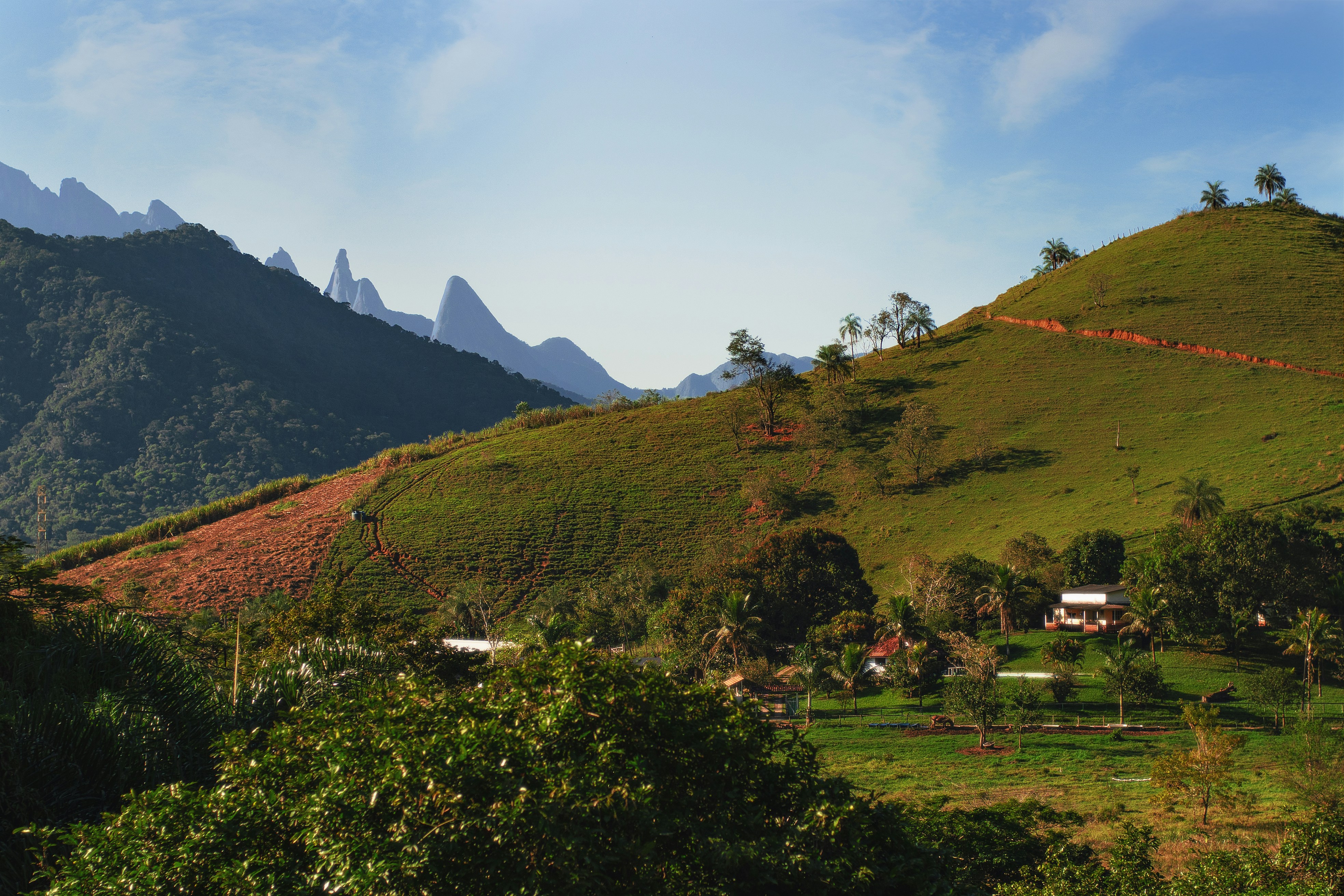 Lush green hills with a small house nestled among trees, framed by distant mountain peaks under a clear sky.