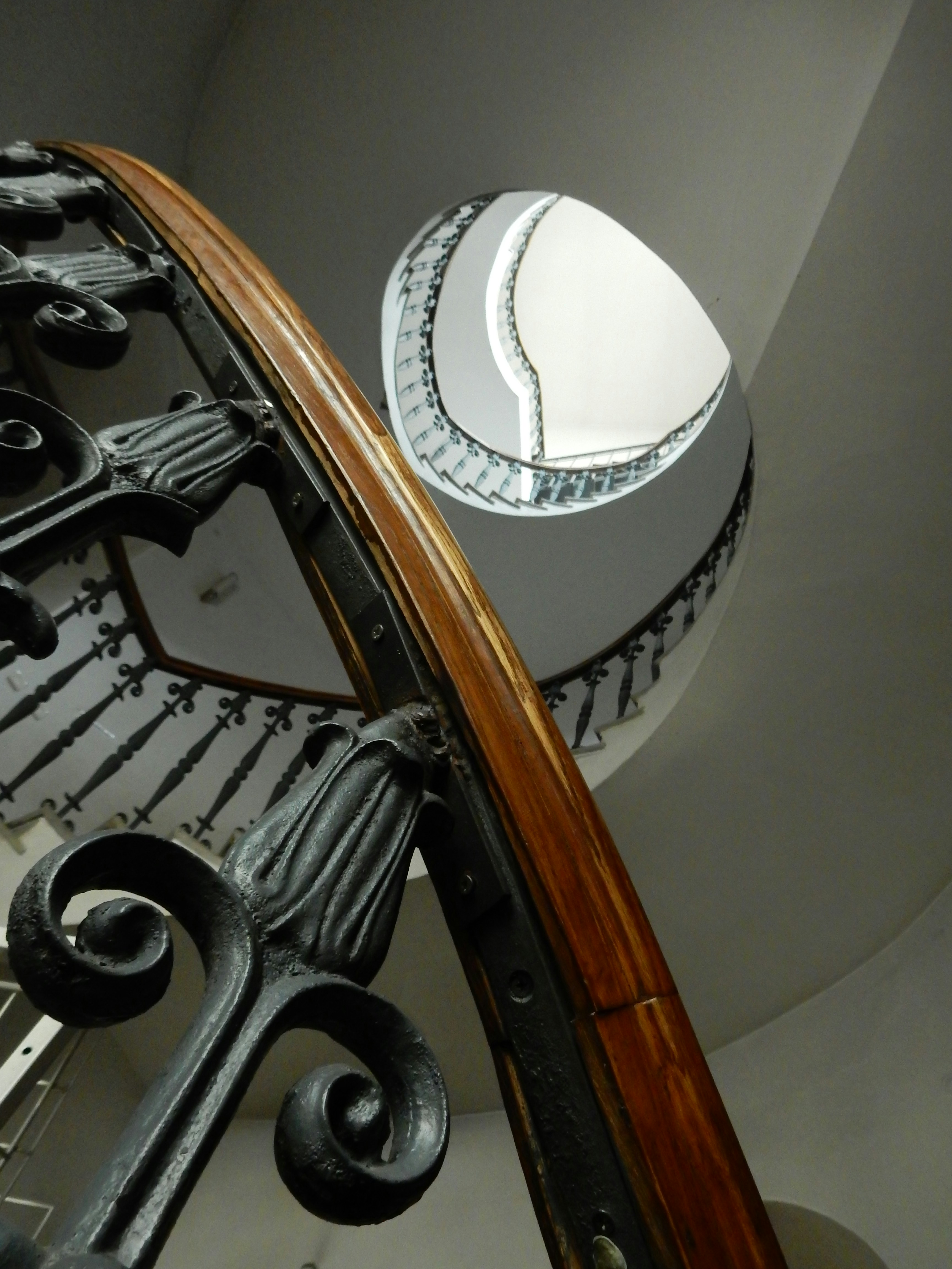 Interior spiral staircase photographed from below, featuring a wooden handrail and decorative iron balusters surrounding a circular opening.