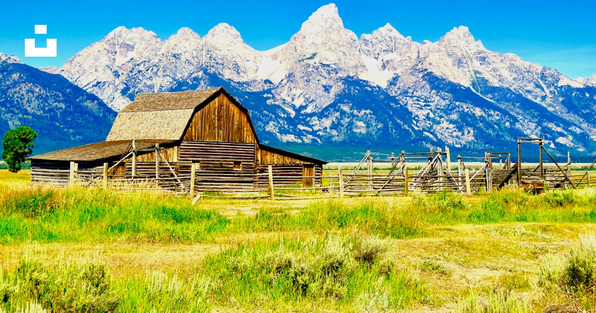 昼間の青空の下の雪に覆われた山の近くの緑の草原に茶色の木造納屋の写真 Unsplashの無料モルモン・ロウ写真