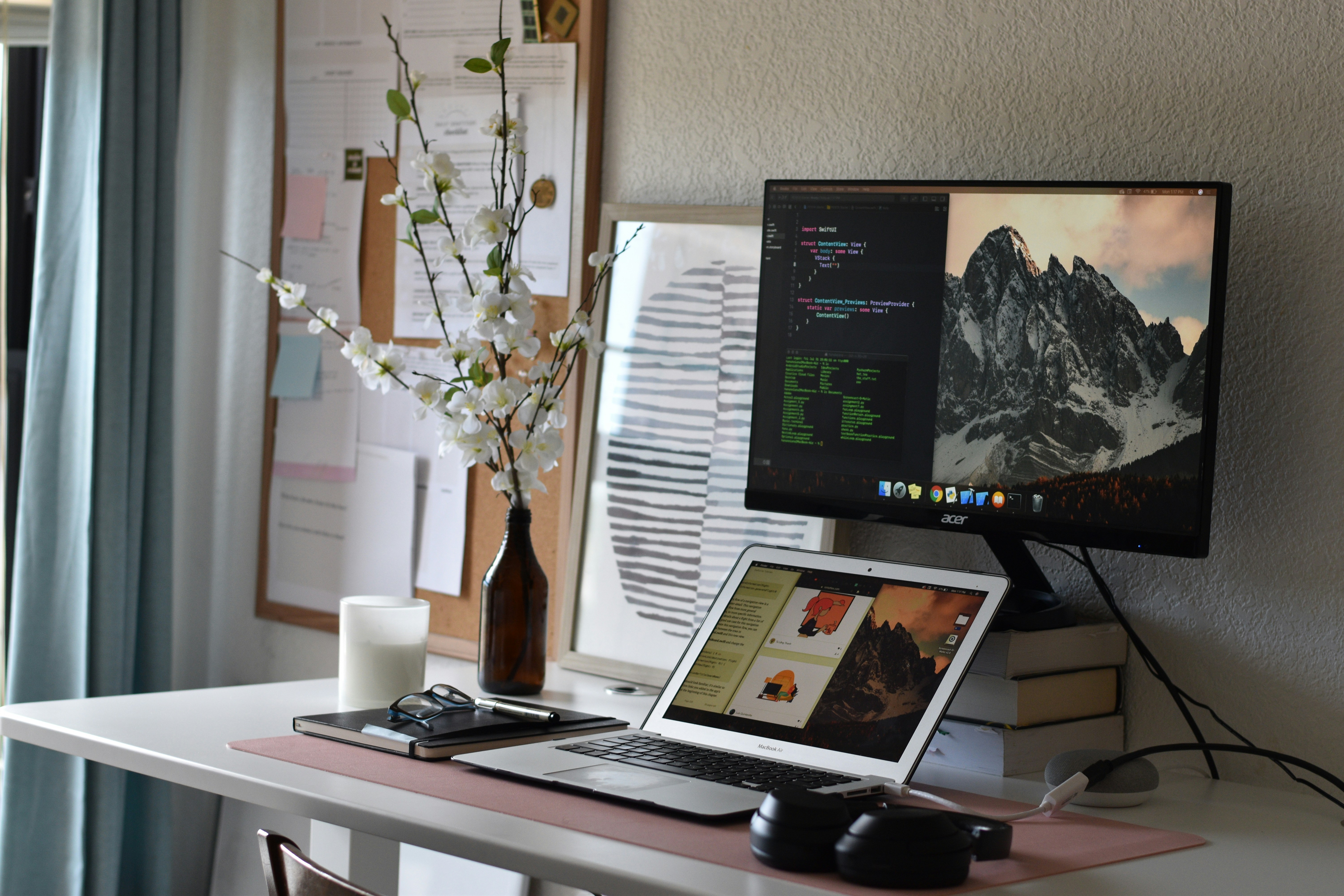 A modern workspace featuring a laptop and a monitor displaying a mountain landscape, complemented by a vase of flowers and a candle. The setting exudes a blend of productivity and tranquility.