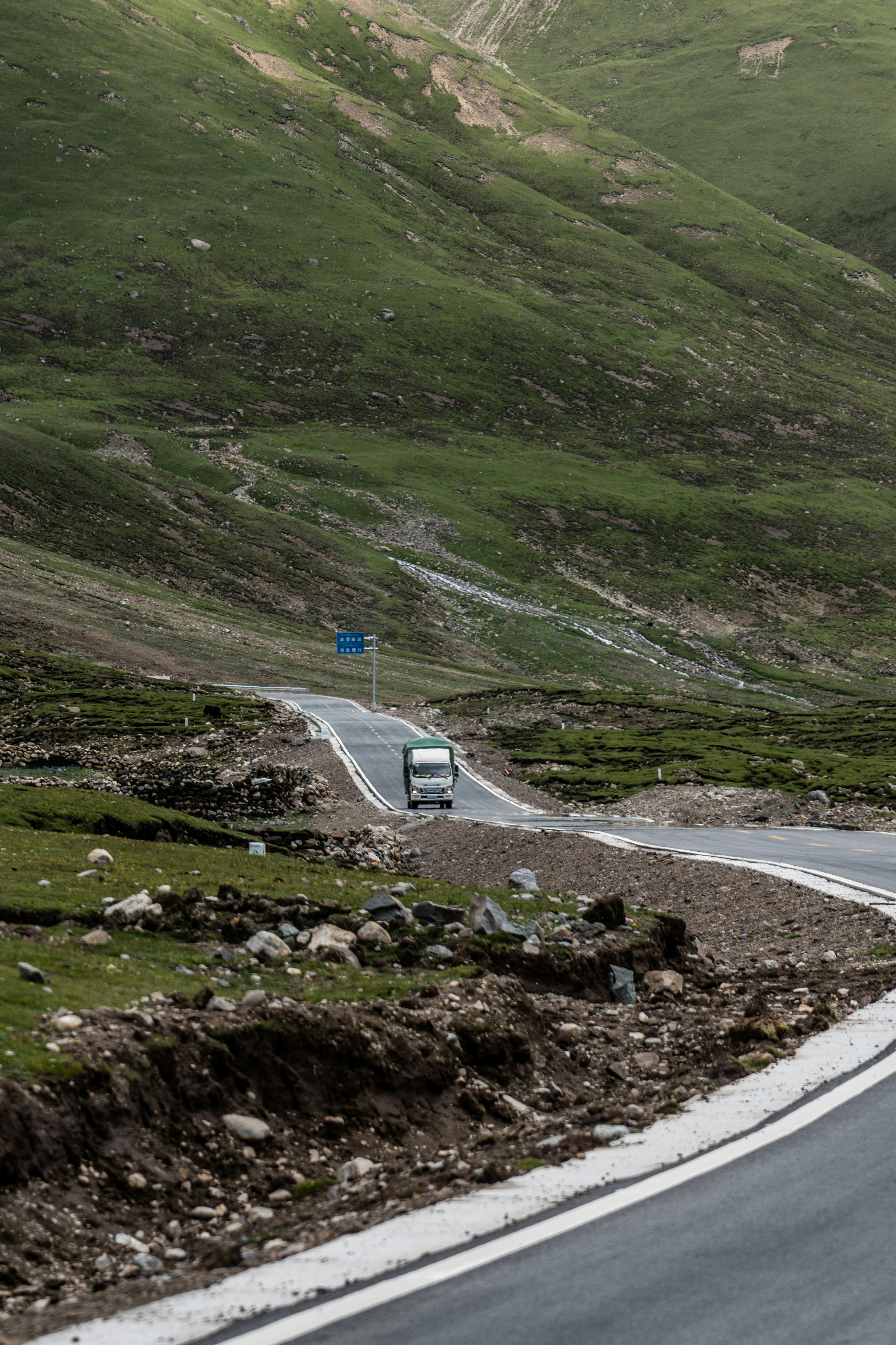 A winding road cuts through lush green hills, with a lone truck navigating the landscape. A road sign stands in the background, marking the route ahead.